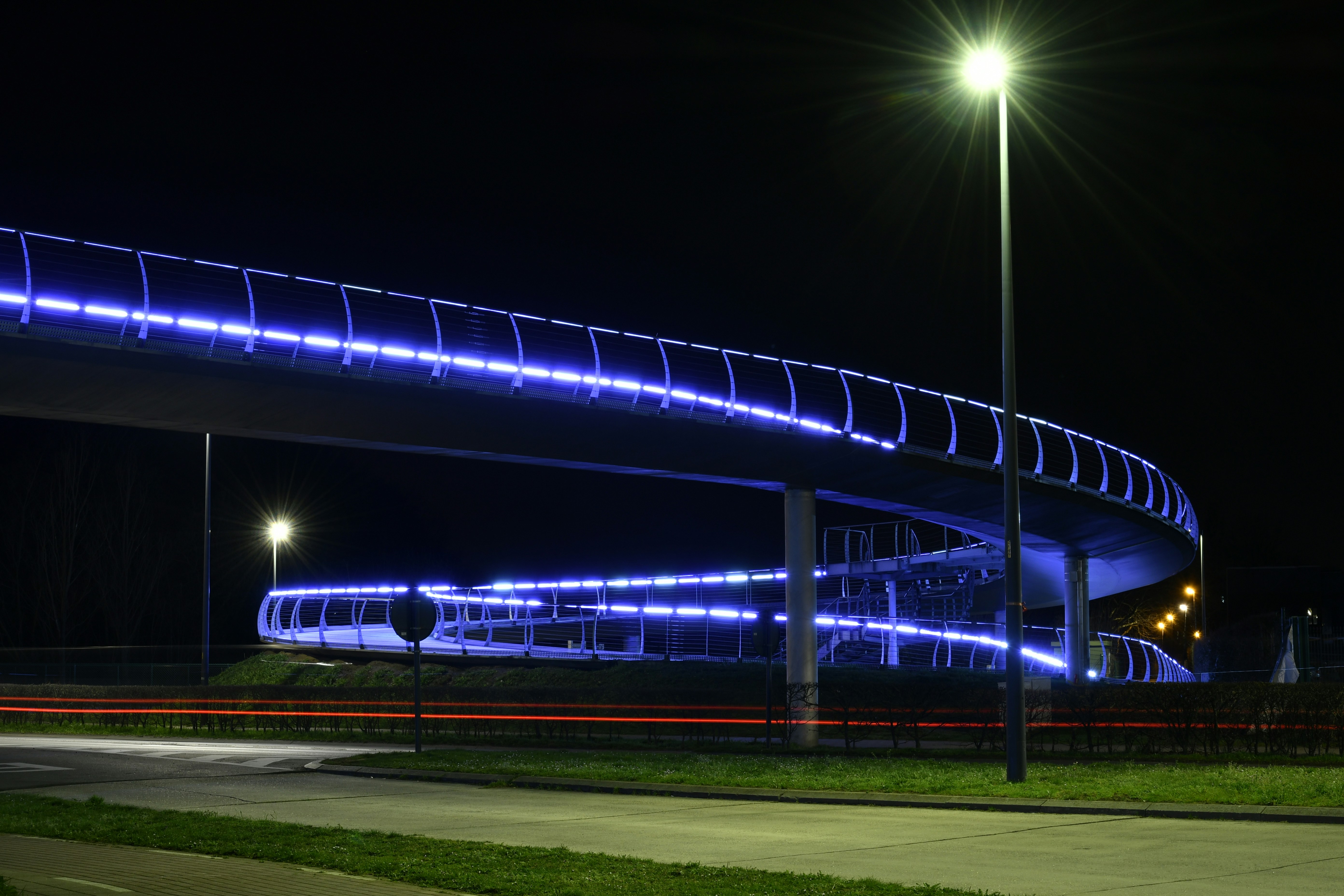 Curved pedestrian bridge illuminated with blue lights at night, creating a striking visual against the dark sky.