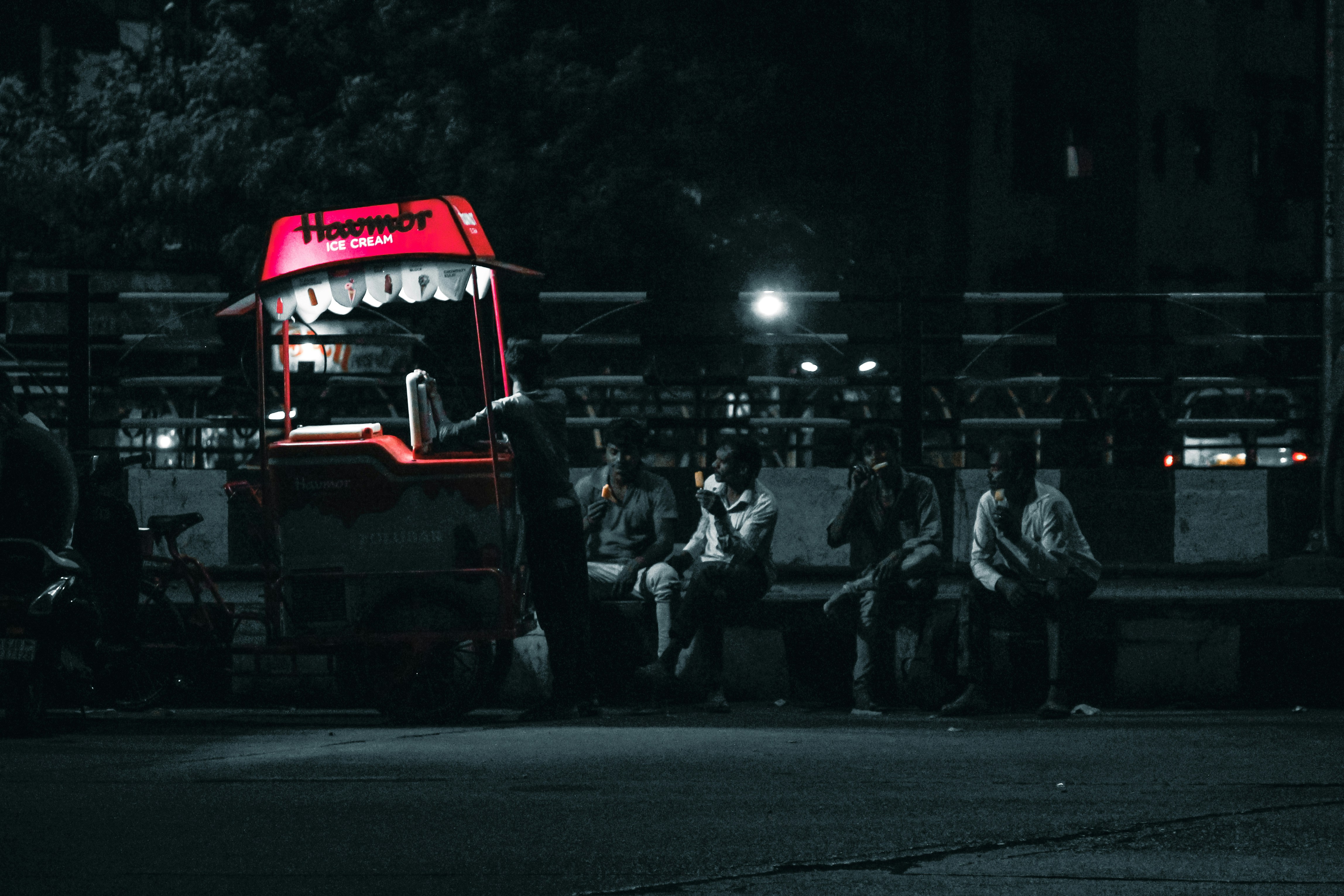 Brightly lit ice cream cart stands out against a dark urban backdrop, with patrons seated nearby enjoying the evening atmosphere.