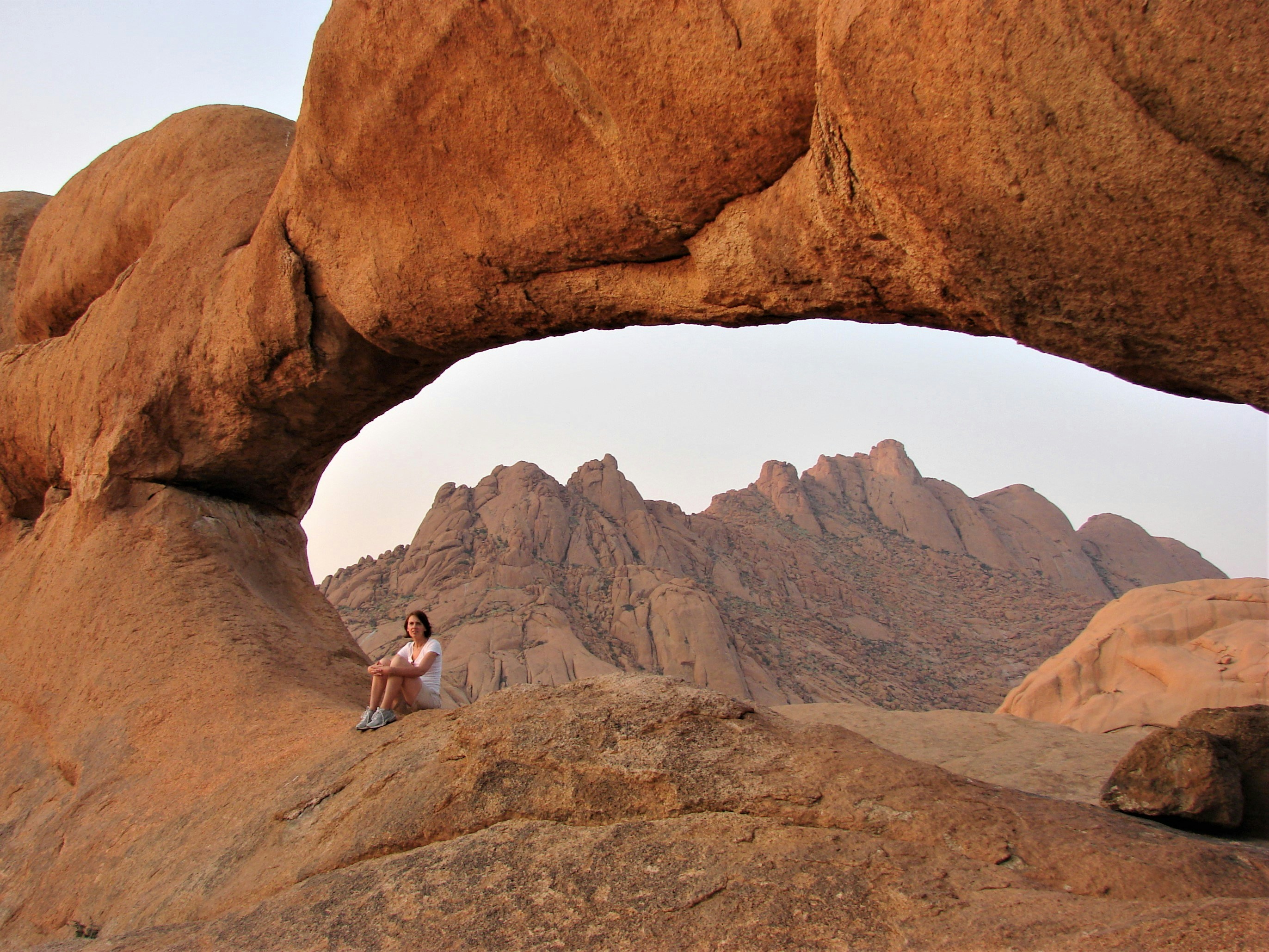 A person sitting peacefully beneath a natural rock arch, surrounded by rugged mountains under a soft sky. The scene captures the harmony between human presence and the grandeur of nature.