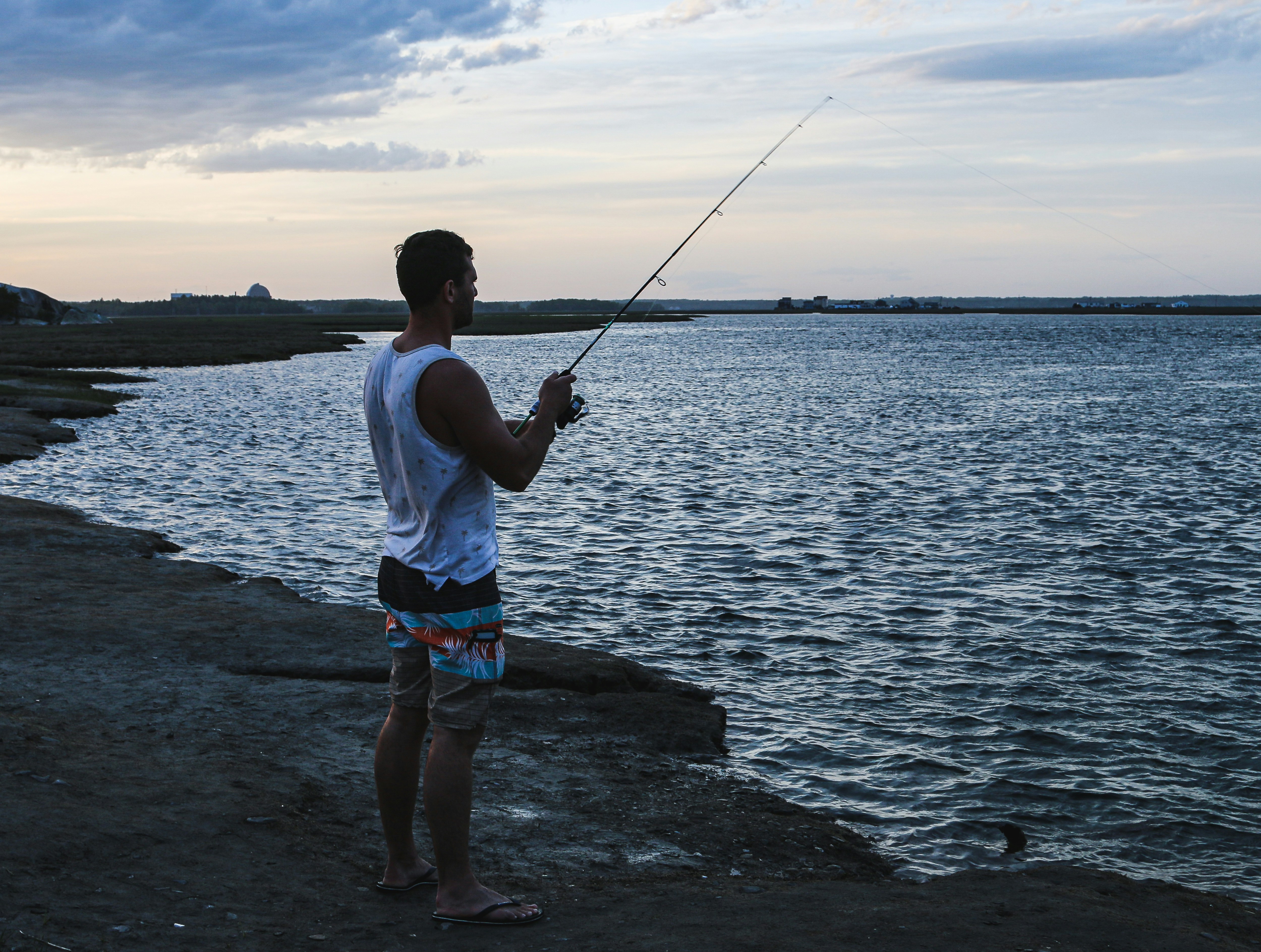 Person fishing along a rocky shoreline at sunset with a calm lake in the background.