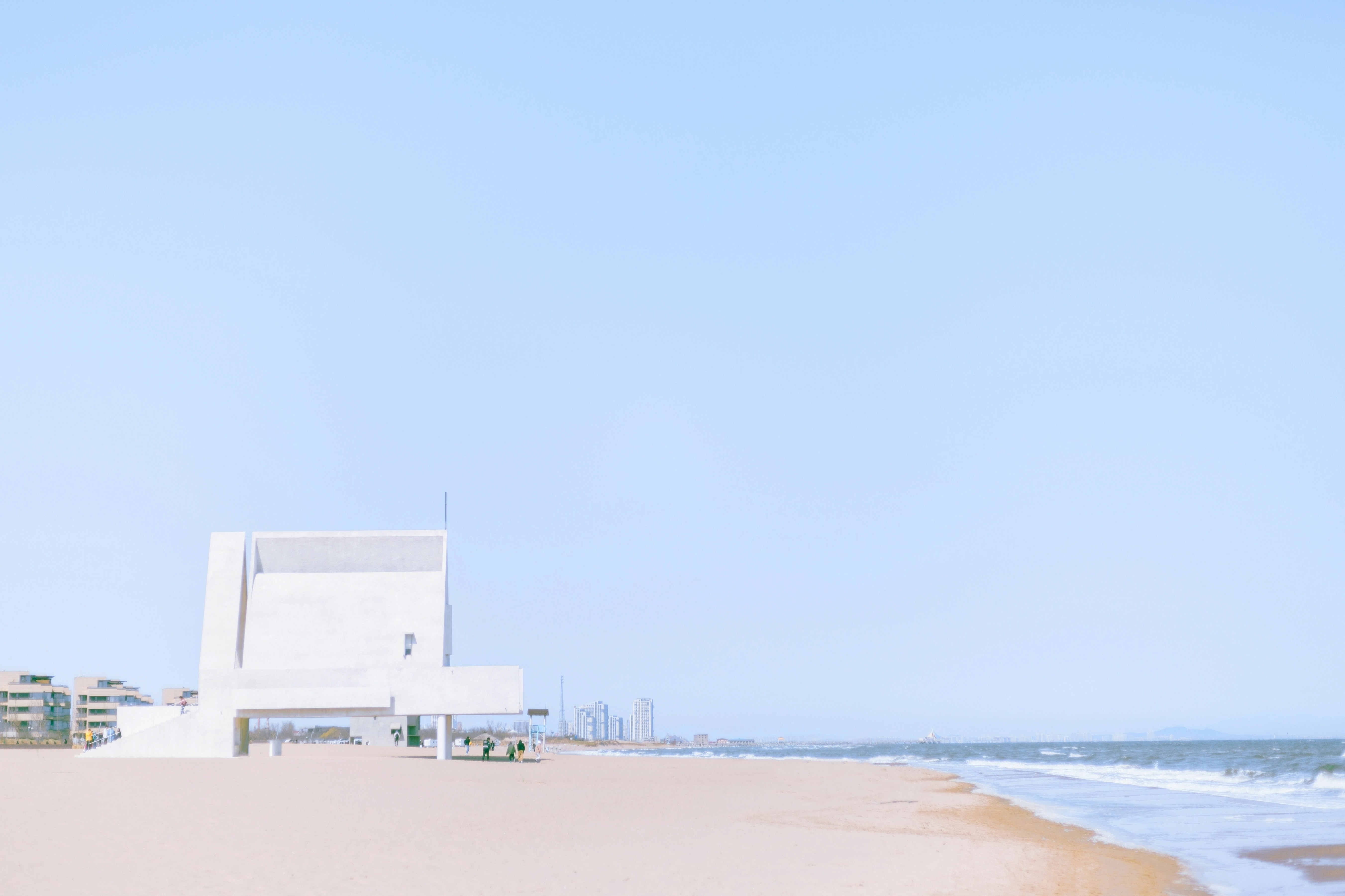 Minimalist architectural structure stands on a sandy beach, framed by a clear blue sky and gentle ocean waves.