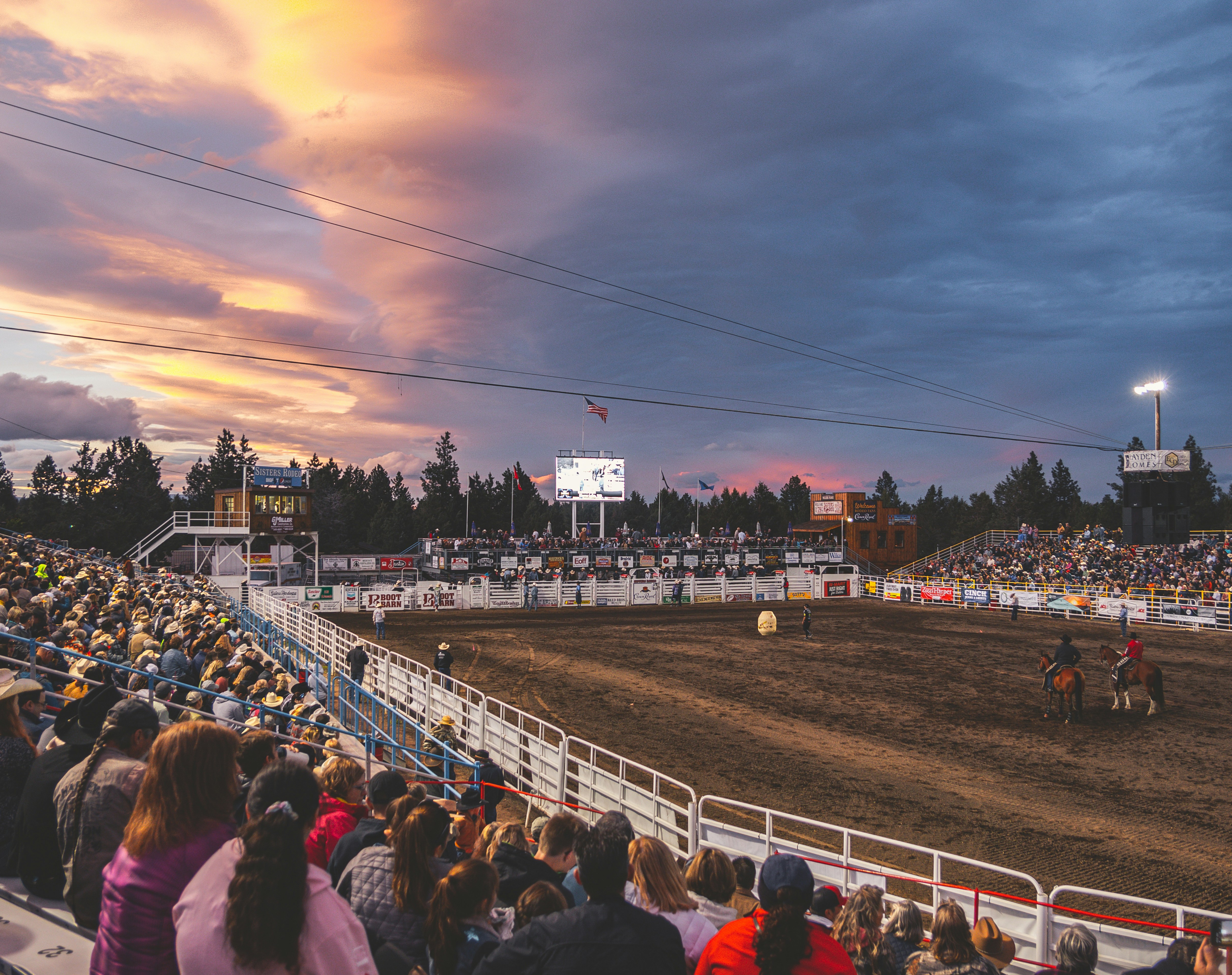 Crowd gathered at a rodeo event under a vibrant sunset, with riders and a bull in the arena. The atmosphere is electric as the evening unfolds.