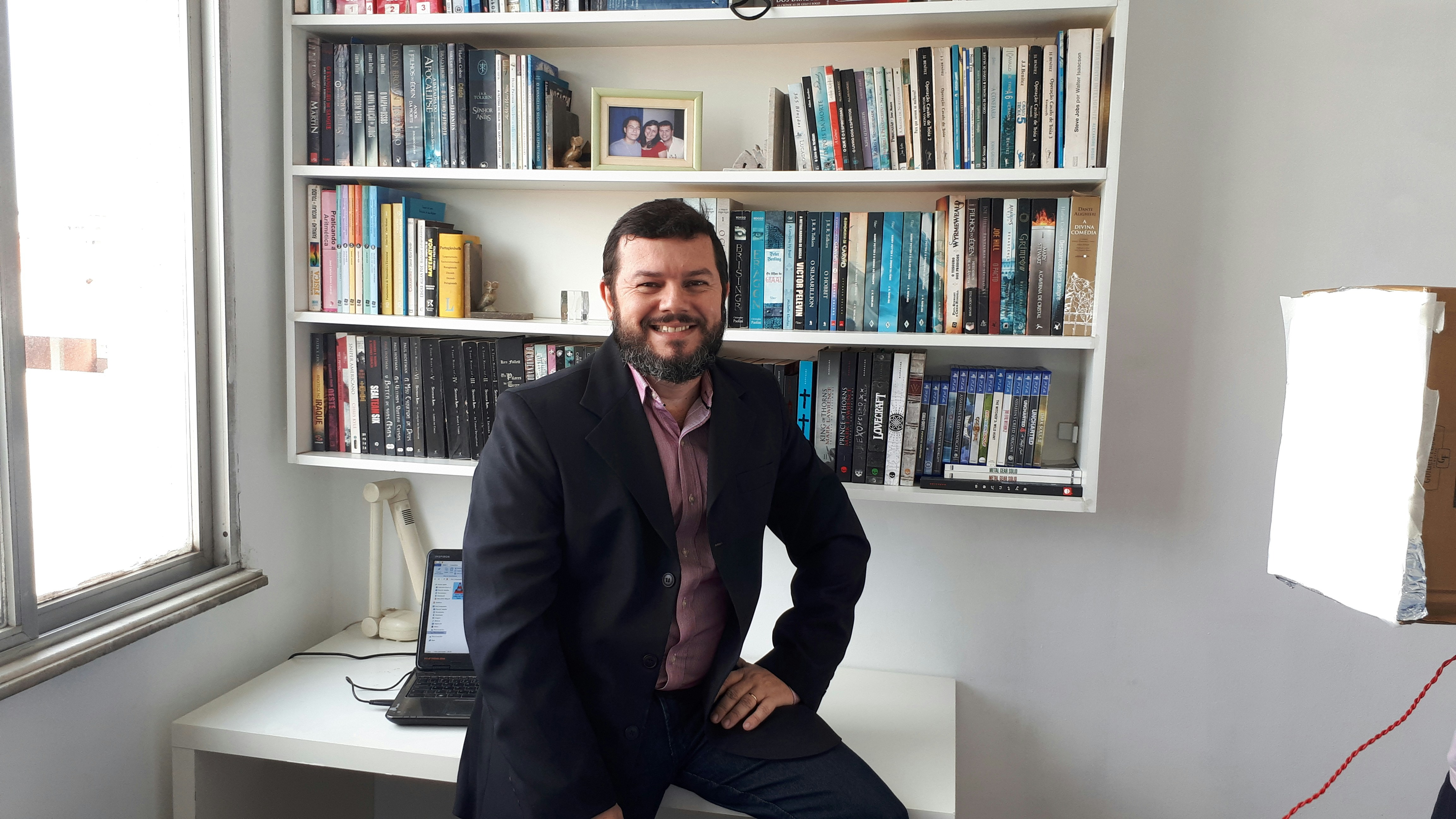 Person sitting at a desk in a home office, surrounded by a wall of bookshelves and a window admitting natural light.