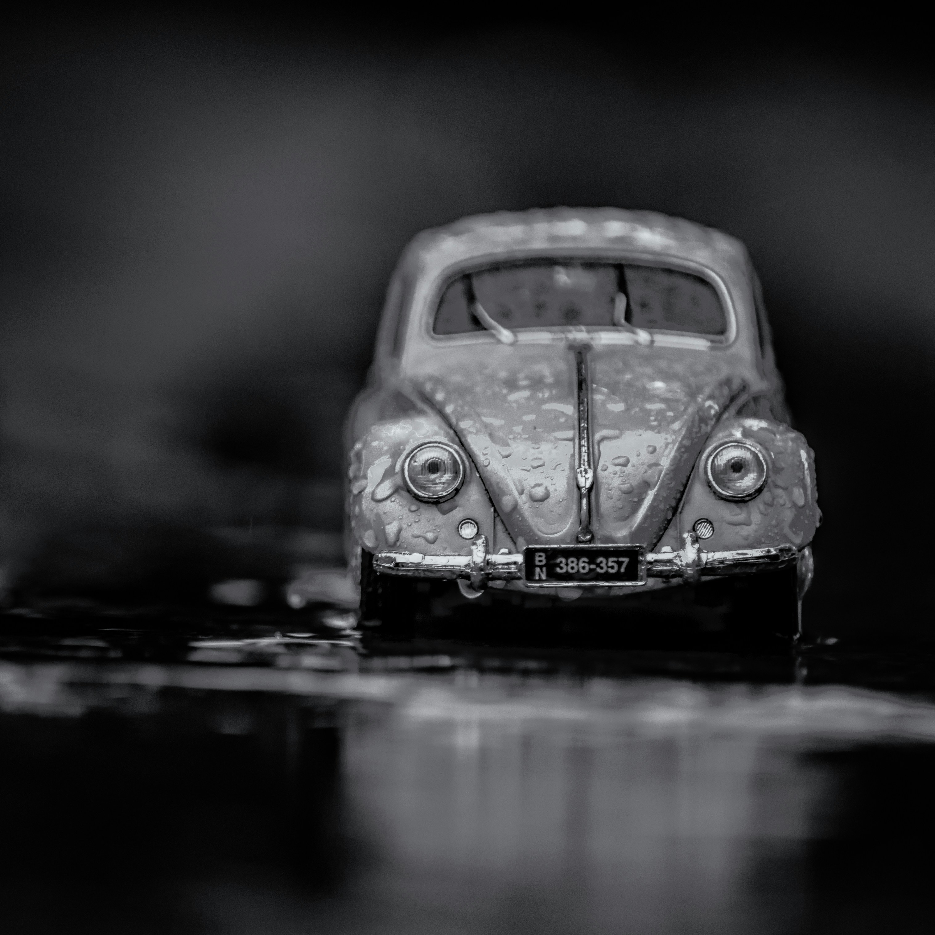 Classic model car glistening with raindrops on a reflective surface in monochrome. The scene evokes nostalgia and tranquility.