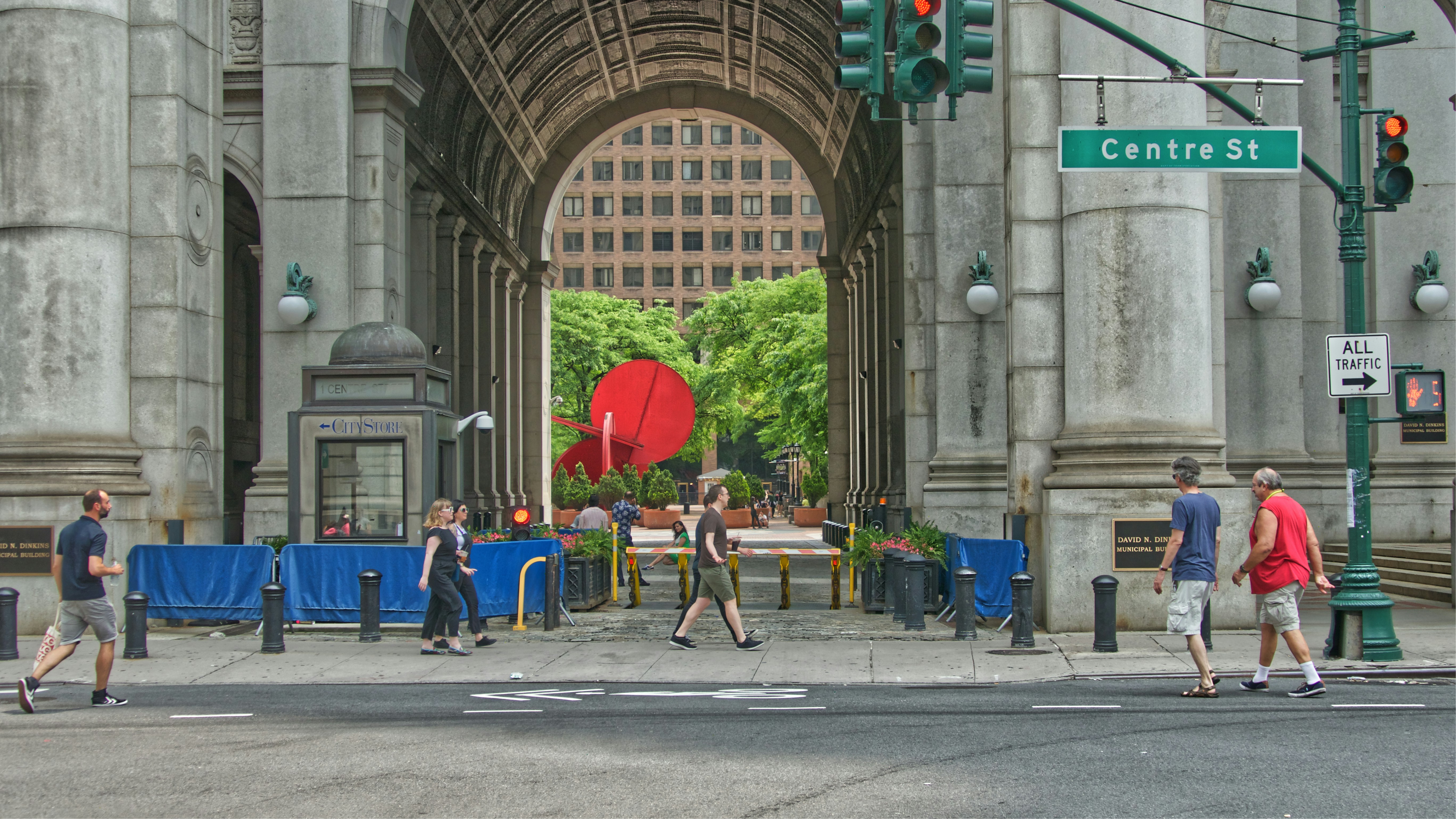 A vibrant red sculpture stands prominently at the end of an architectural archway, framed by bustling pedestrians and lush greenery.
