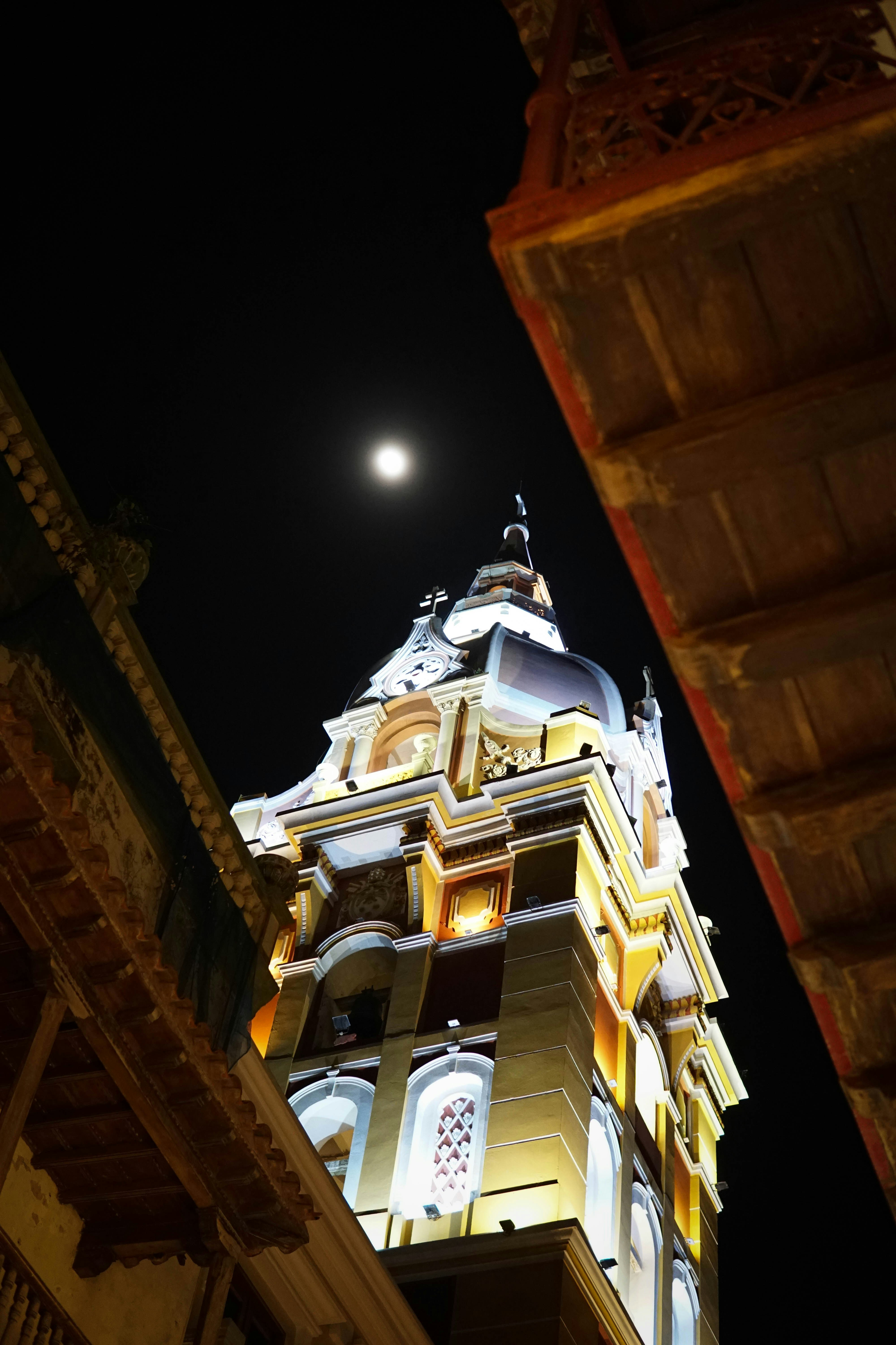 Illuminated tower against a night sky with the moon, framed by surrounding architecture.