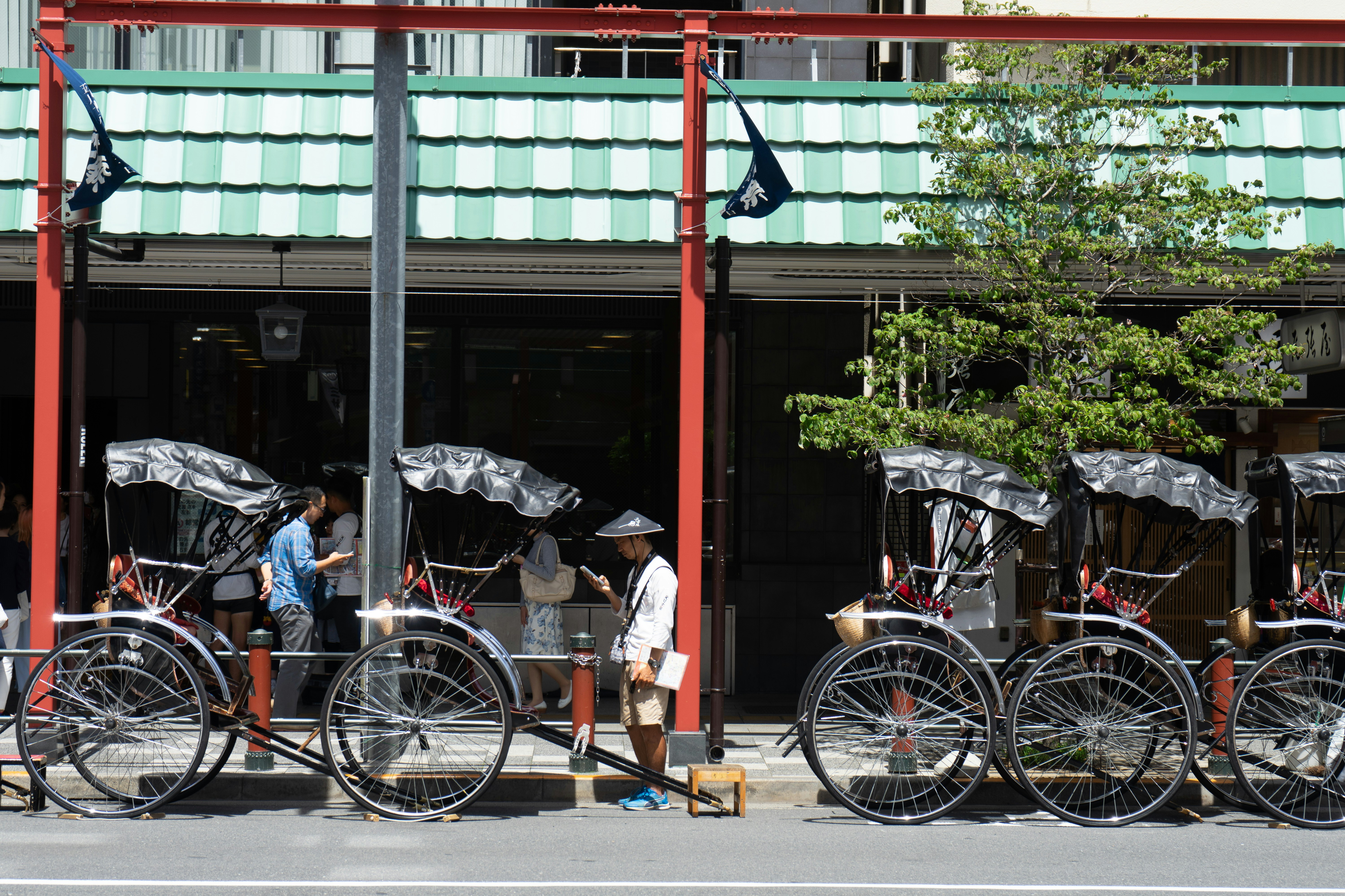 A row of traditional rickshaws lined up along a bustling street, with pedestrians engaging in lively conversation near a shaded storefront.