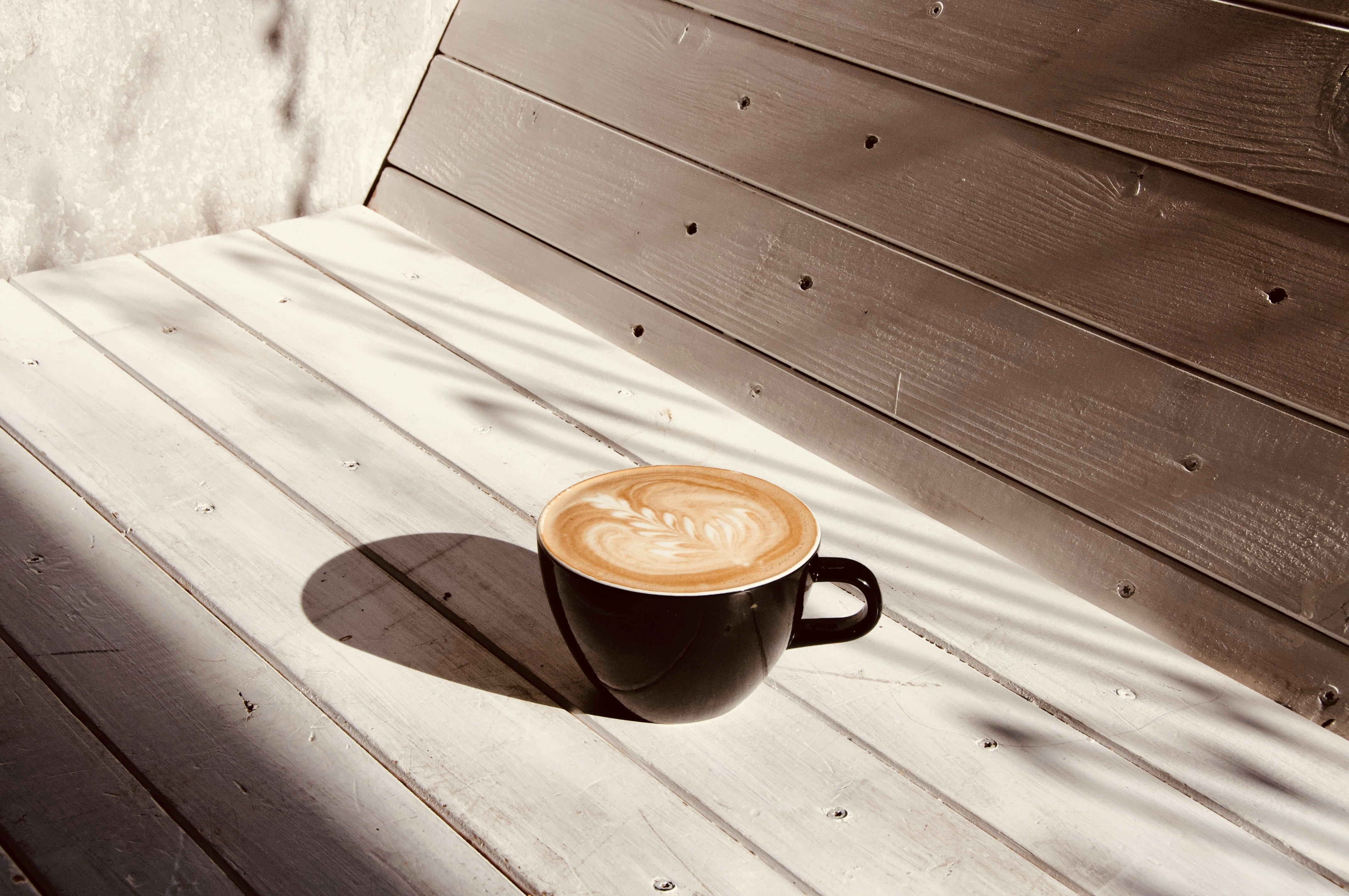 Artfully crafted latte in a black cup resting on a sunlit wooden surface, surrounded by soft shadows.