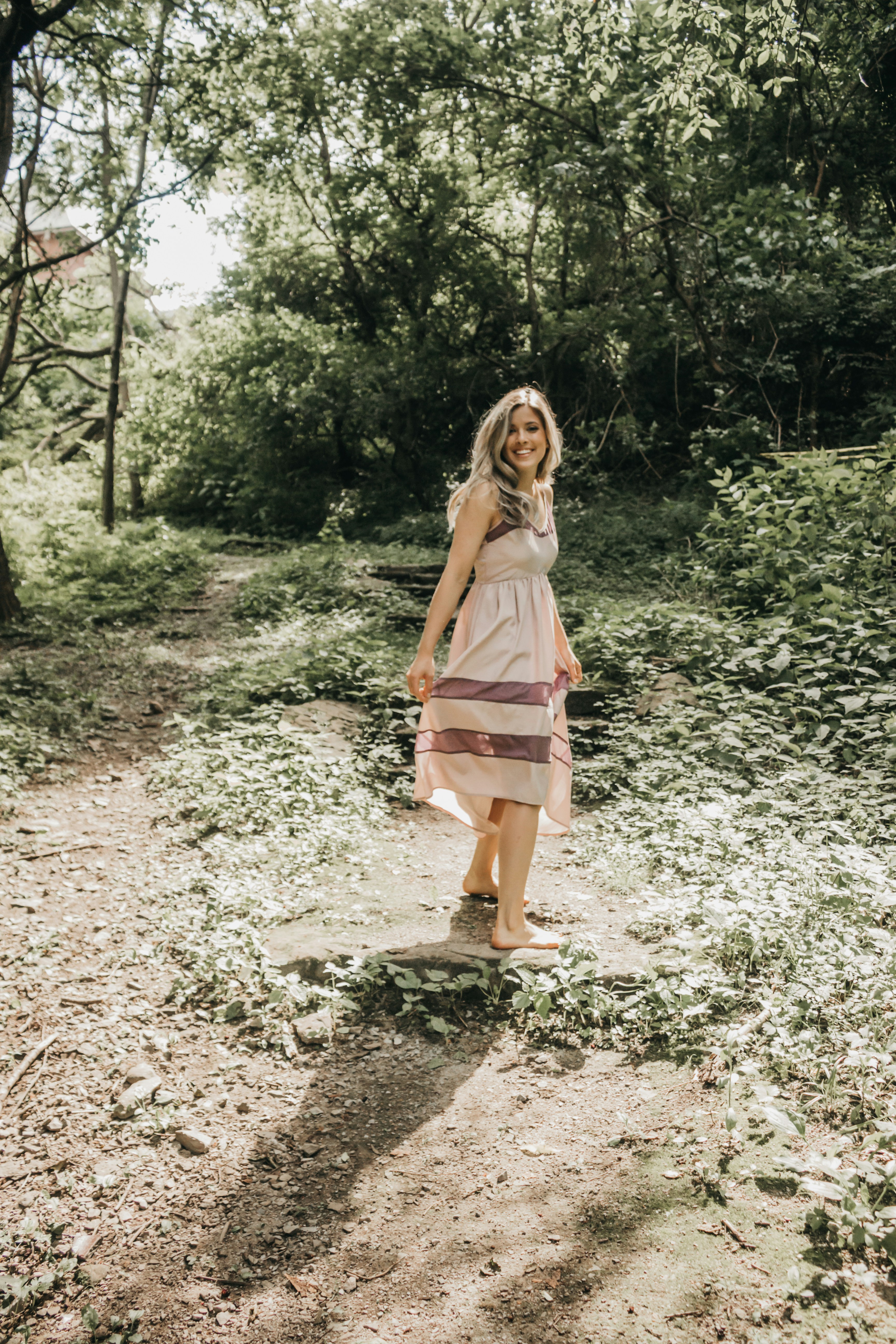Woman in a flowing beige dress walking barefoot on a forest path under dappled sunlight.