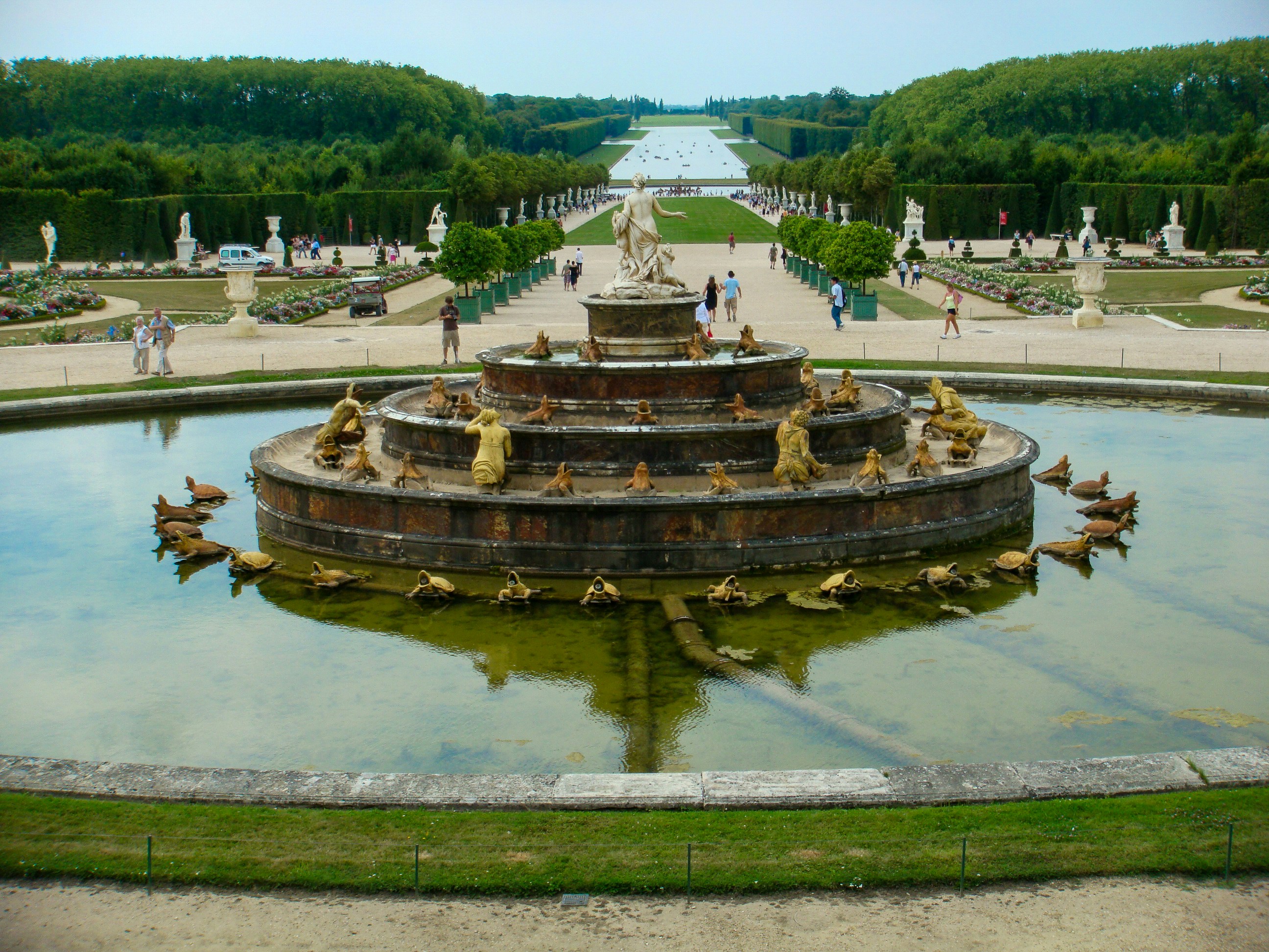 Ornate tiered fountain with golden sculptures in a formal garden, framed by lush greenery and a distant canal.