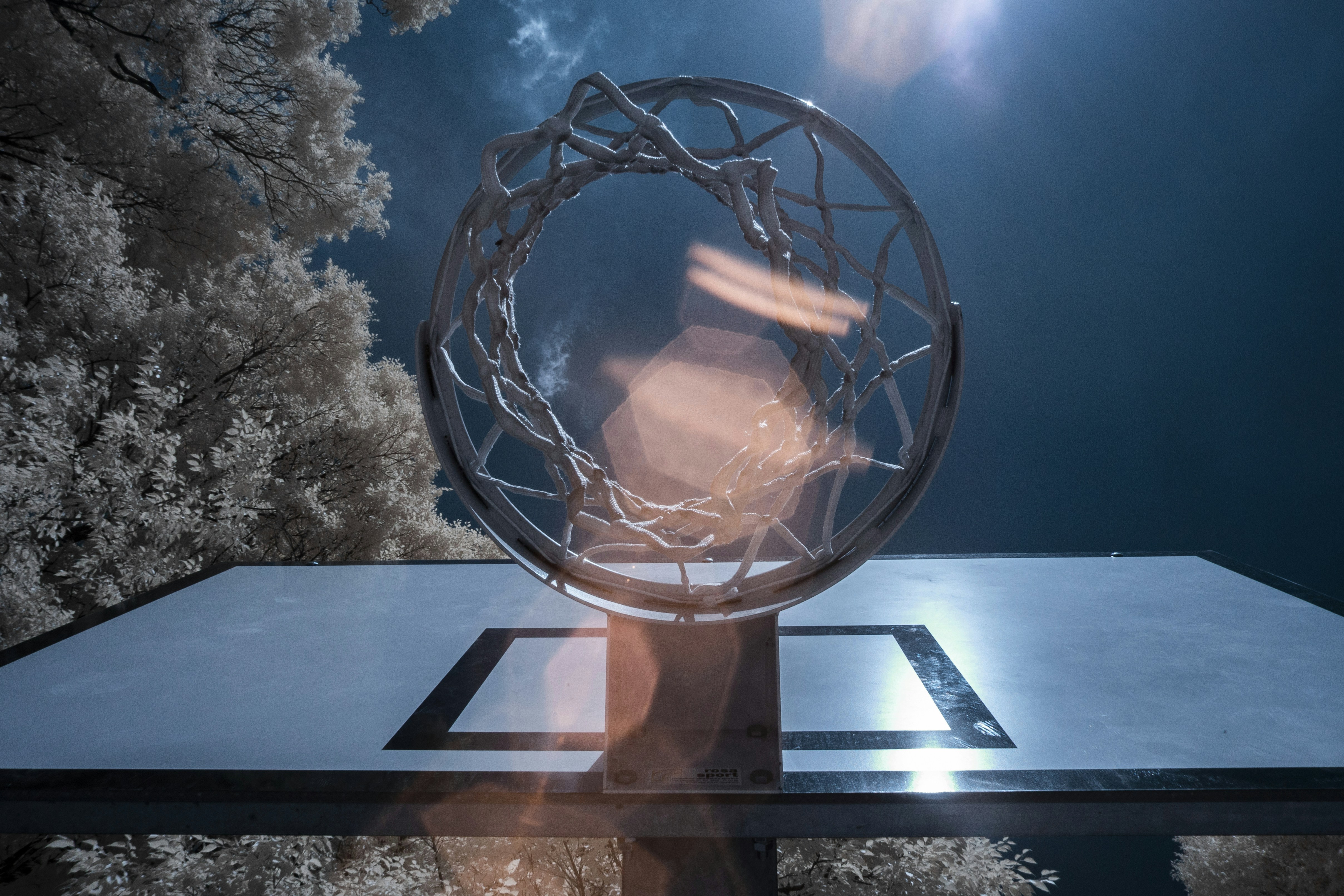 Low-angle photograph of a basketball hoop and backboard set against a deep blue sky, with an orange motion blur passing through the hoop.
