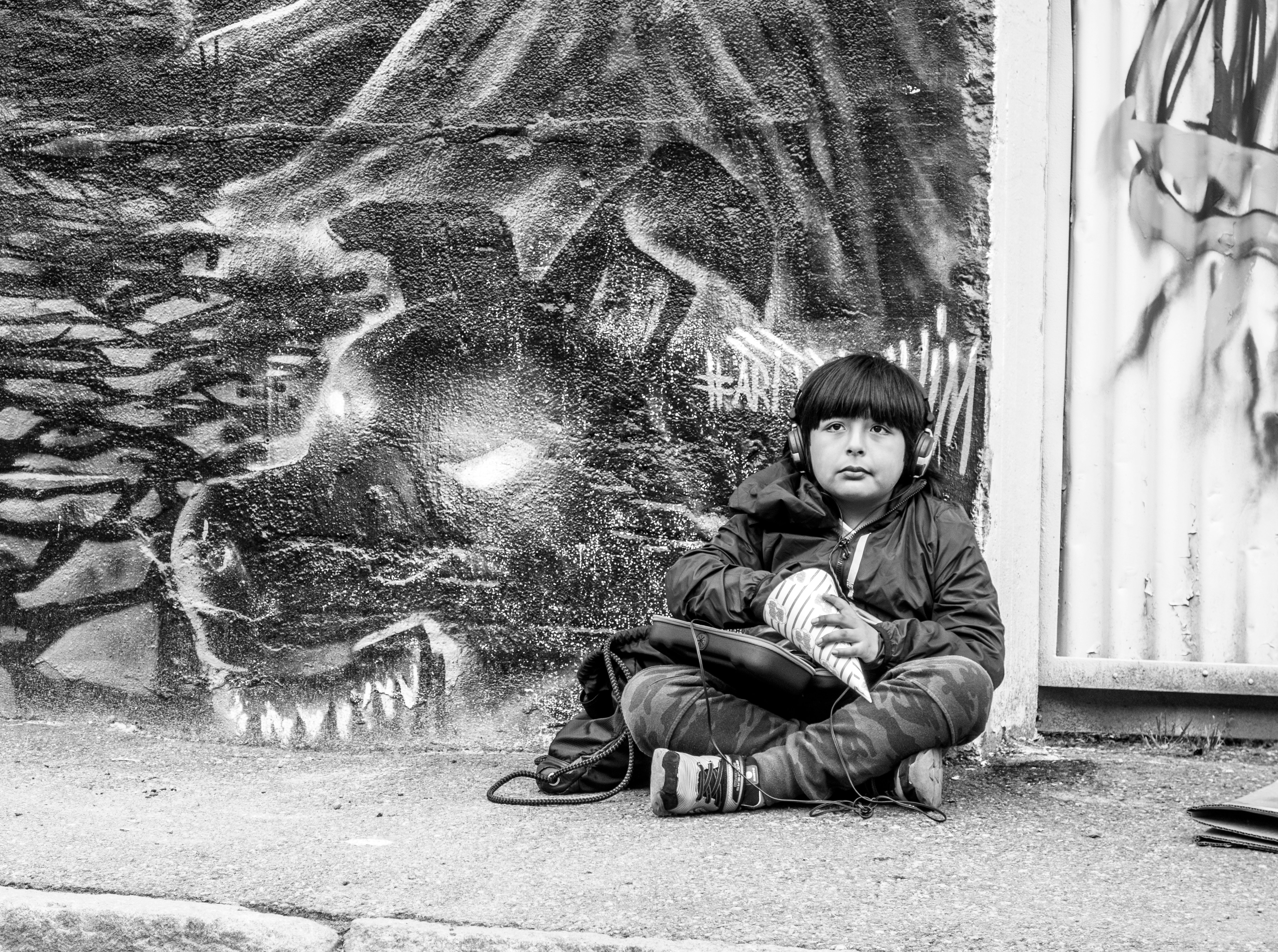 A young boy sits against a vibrant mural, lost in thought, with headphones on, embodying the intersection of childhood and urban life.