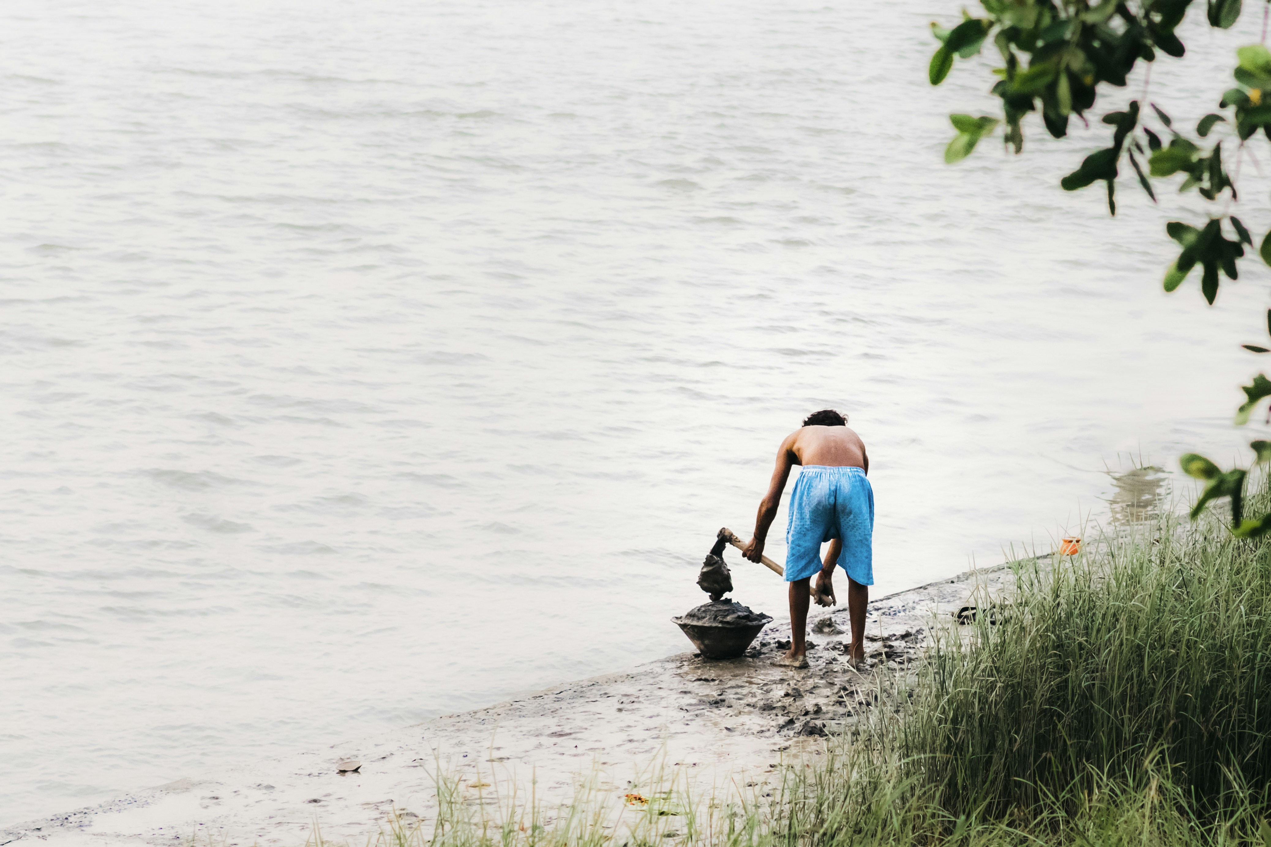 Person in blue shorts working with a draw hoe on a sandy shoreline near calm water, surrounded by greenery.