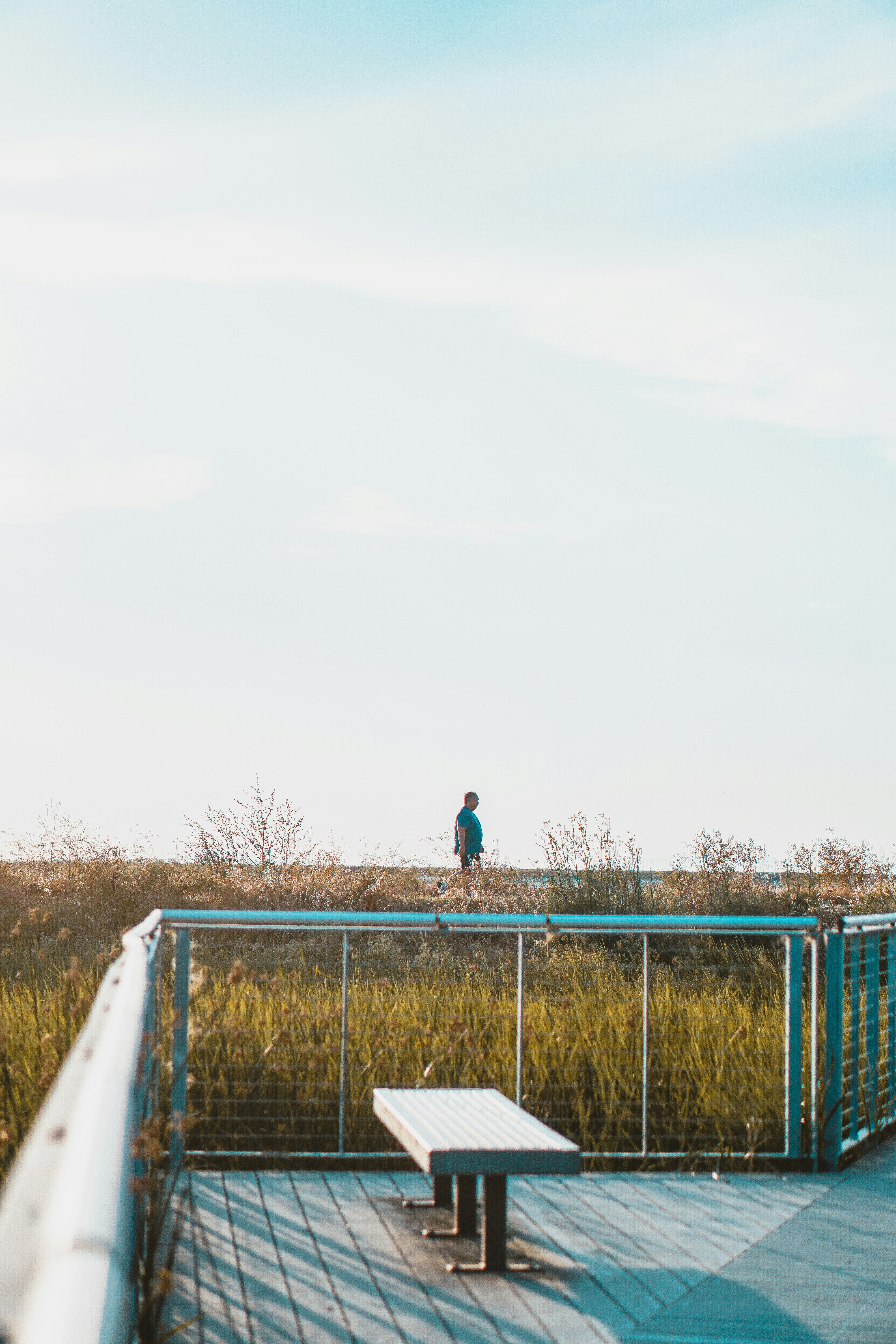 A lone figure walking along a grassy path under a clear sky, framed by a wooden deck and railing. The scene evokes a sense of tranquility.