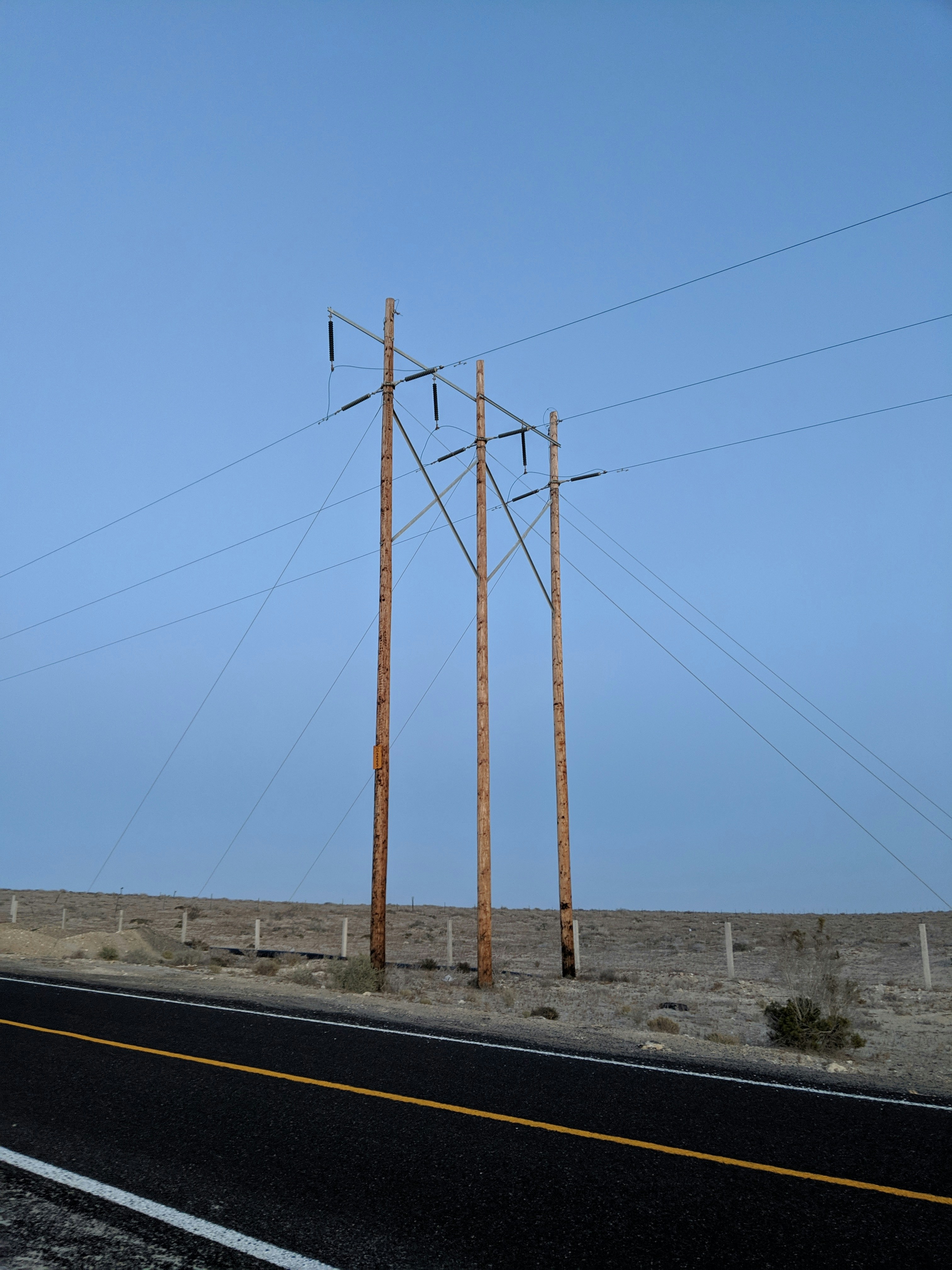 Desert landscape photograph featuring three tall wooden power poles with guy wires against a clear blue sky. A paved road and fence line the arid foreground.