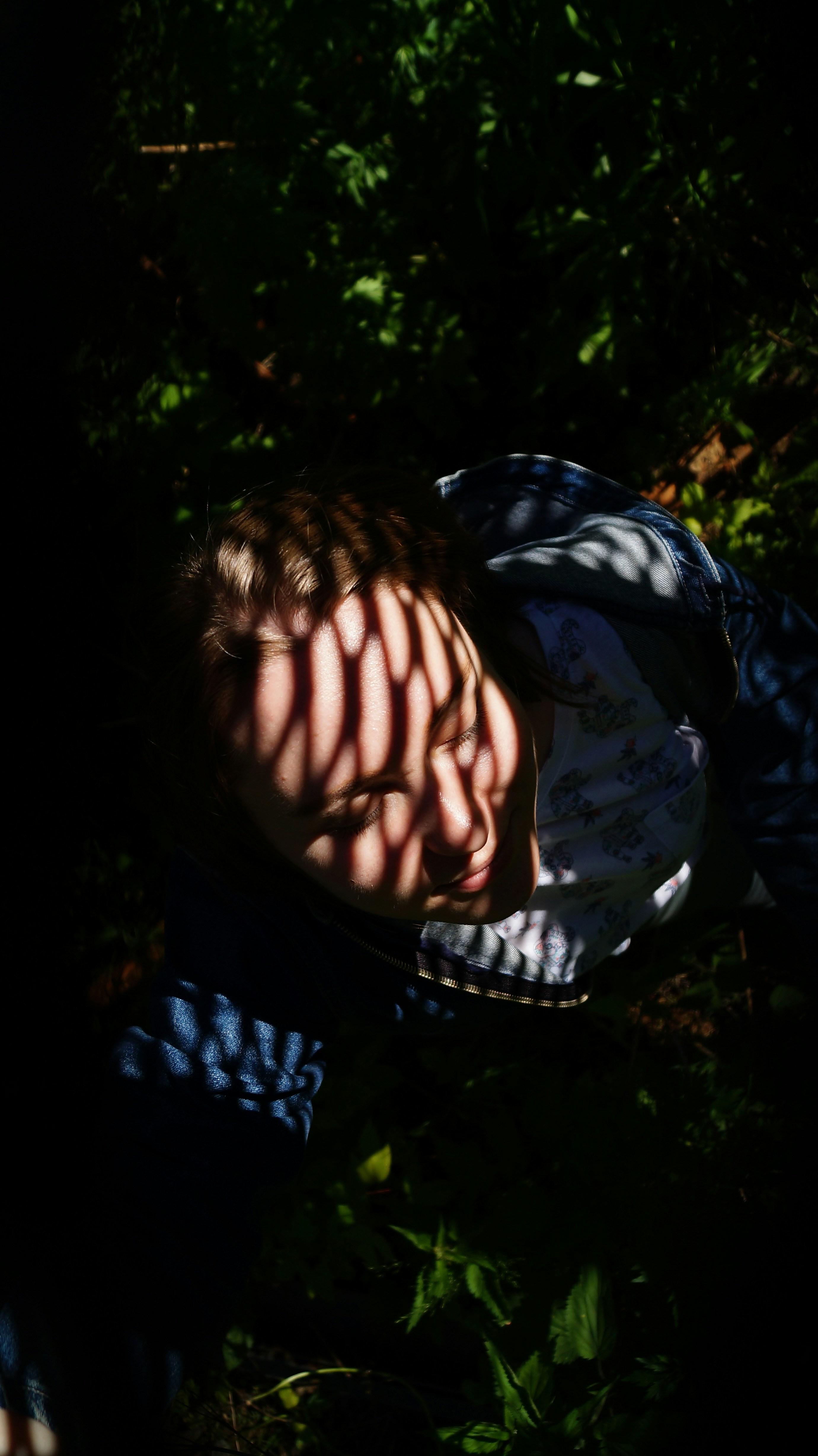 A person surrounded by lush greenery, illuminated by dappled sunlight filtering through a patterned shadow. The interplay of light and shadow creates a striking visual effect.