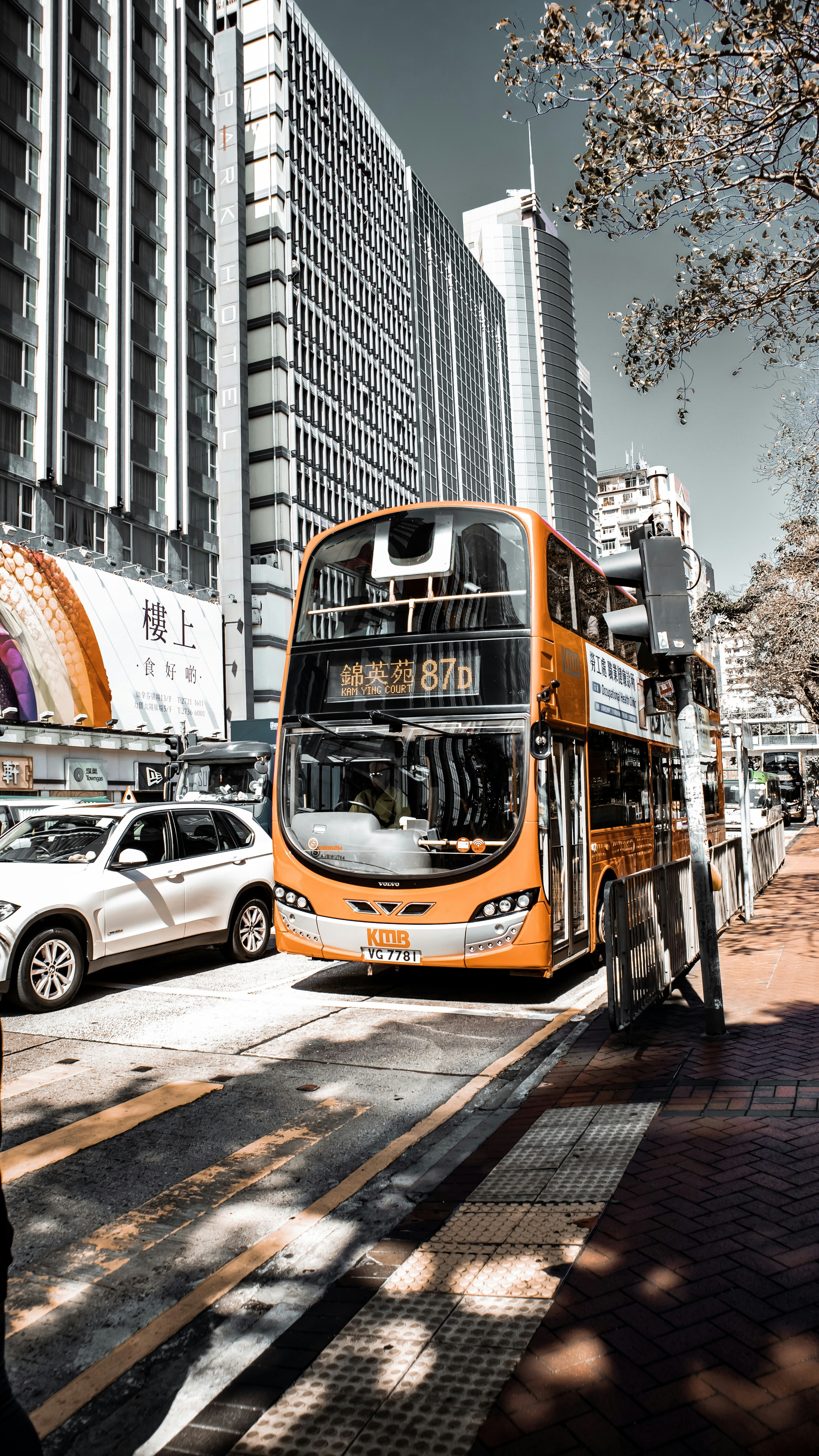 Amber double-decker bus threads a sunlit urban canyon between glass towers