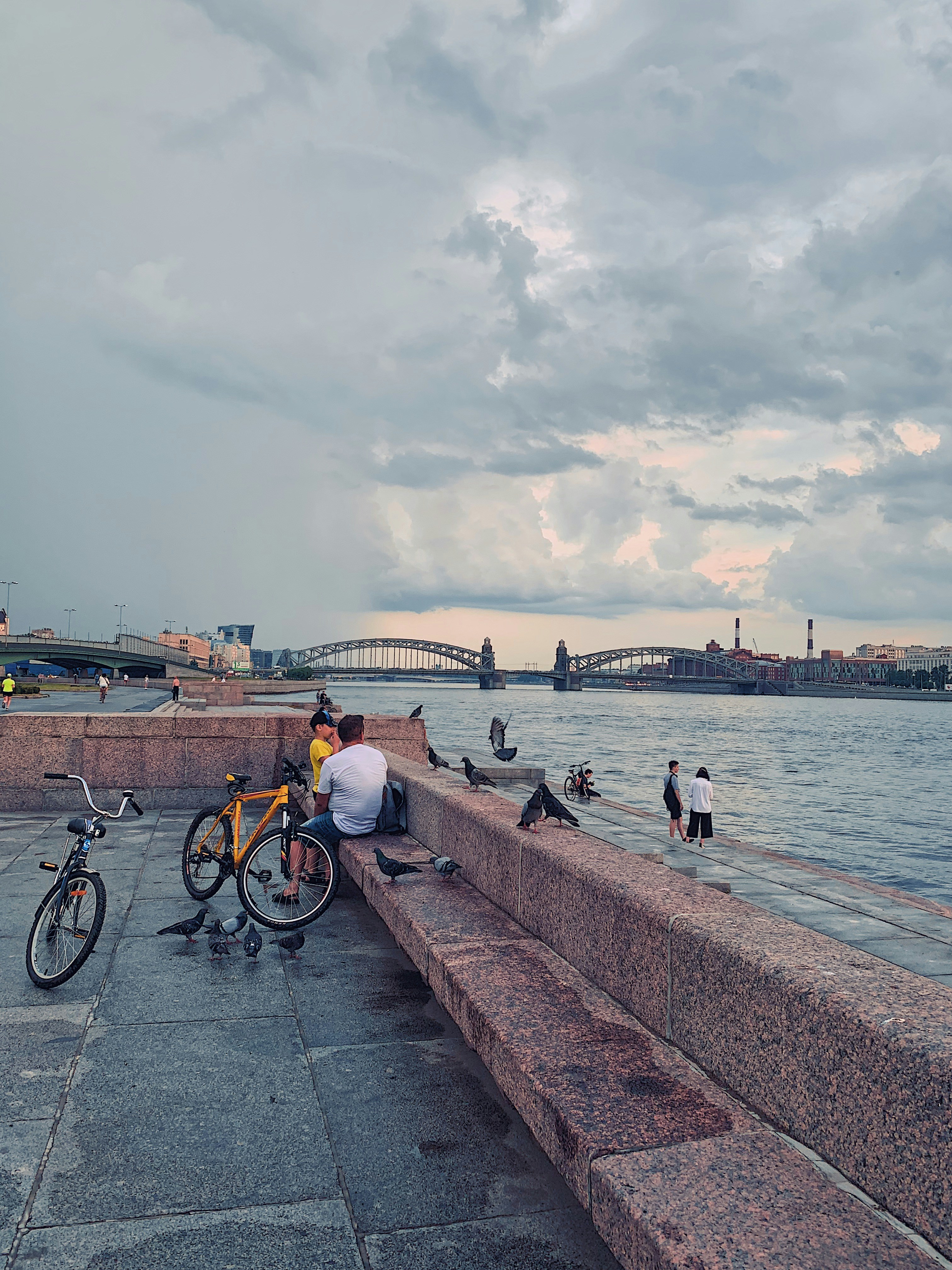 Riverside Pause: Bicycles, People, and a Distant Arch Bridge