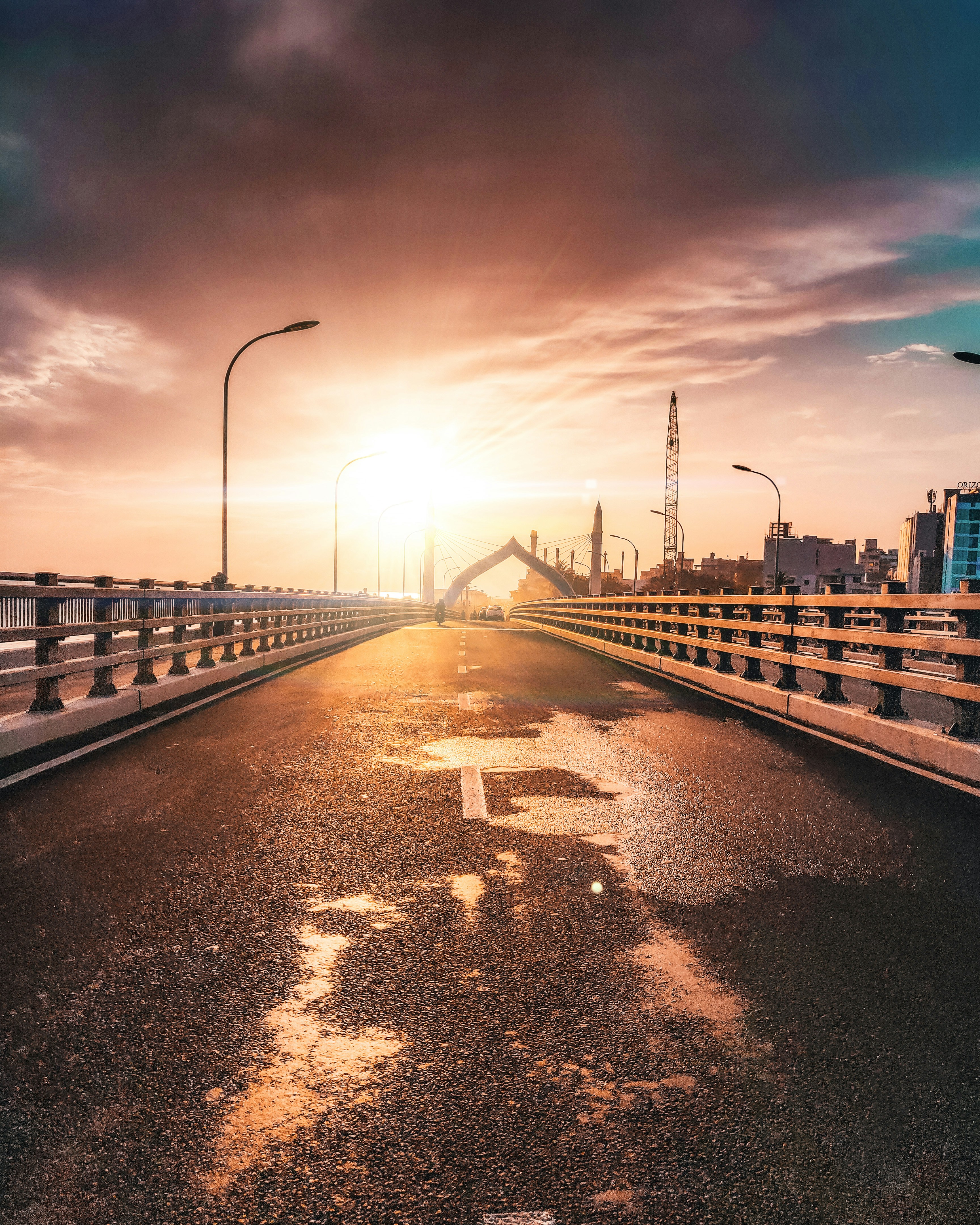 A serene urban bridge bathed in warm sunset hues, with reflective wet pavement leading towards a glowing horizon.