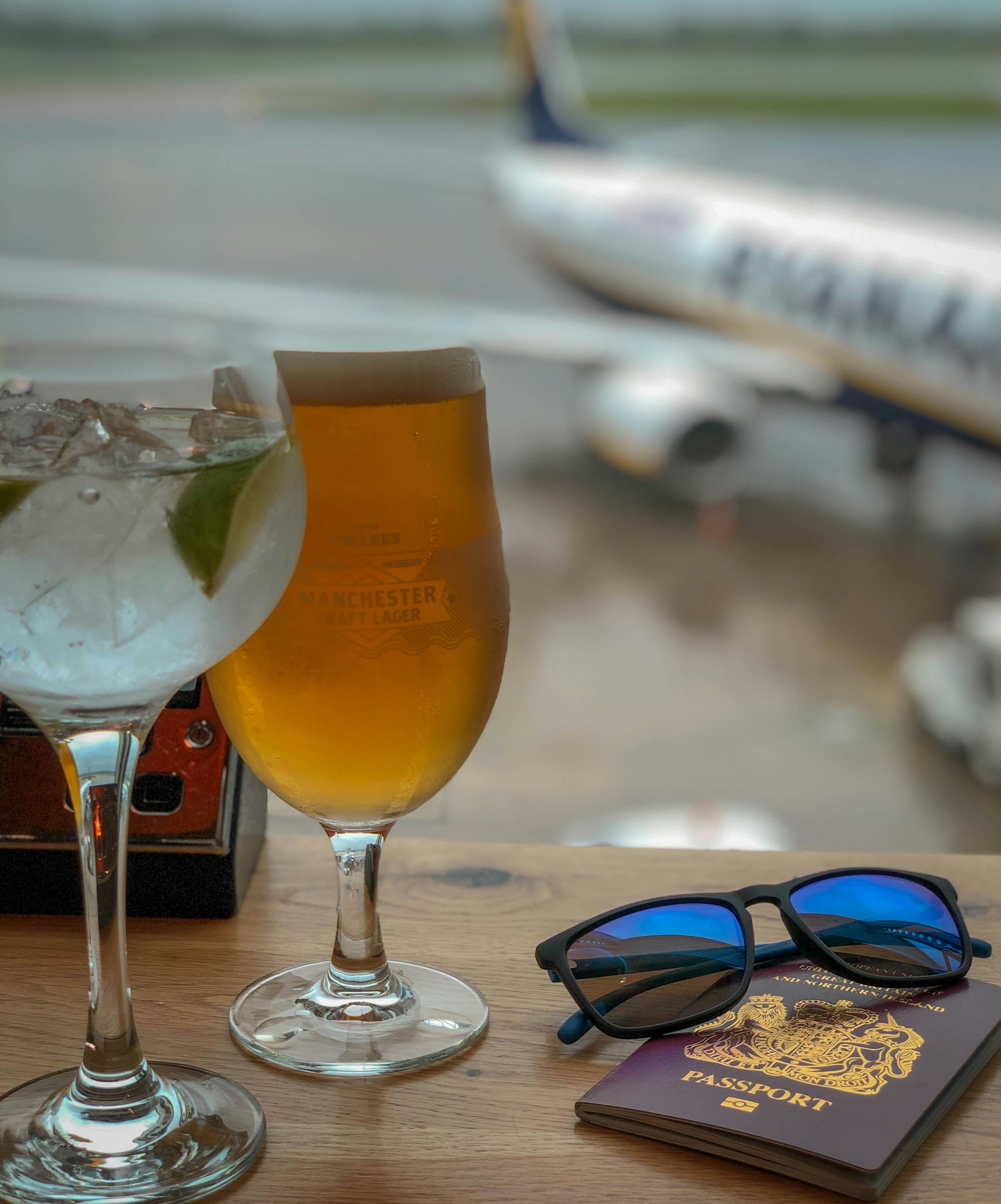 Two drinks sit on a wooden table with a passport and sunglasses nearby, while a Ryanair airplane is blurred in the background.
