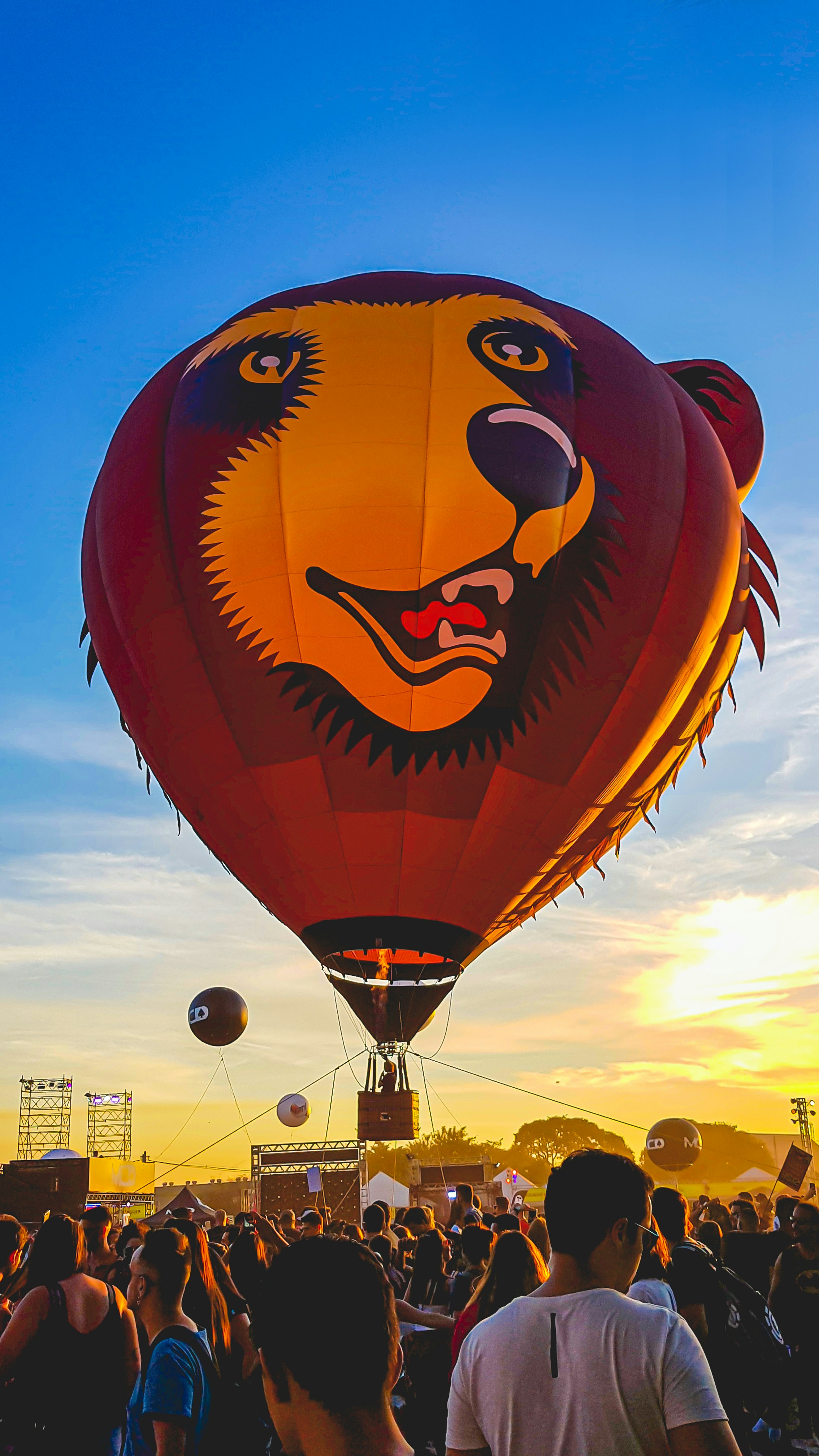 Colorful hot air balloon shaped like a bear rises above a lively crowd during a festival at sunset.