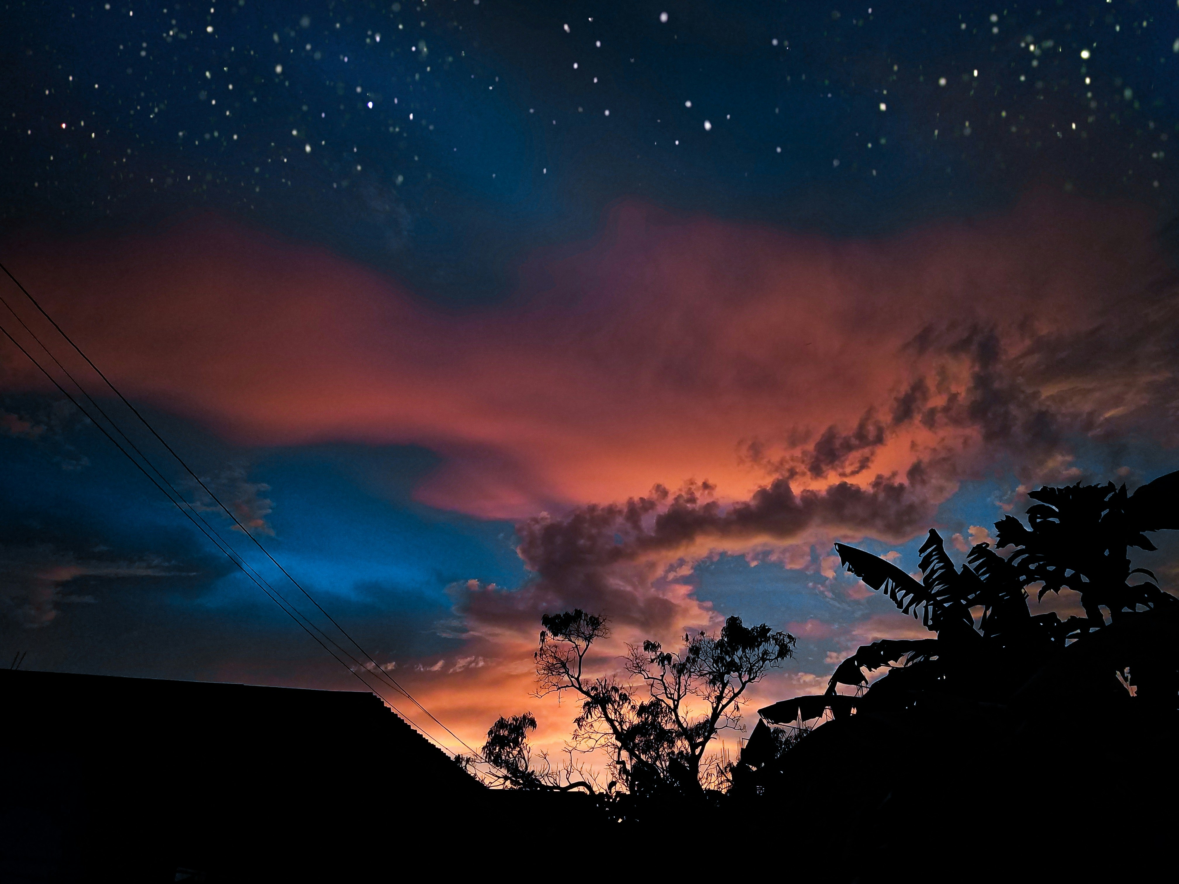 Vibrant sunset hues blend with emerging stars, silhouetting trees and rooftops against a dramatic sky. The scene captures the transition from day to night.