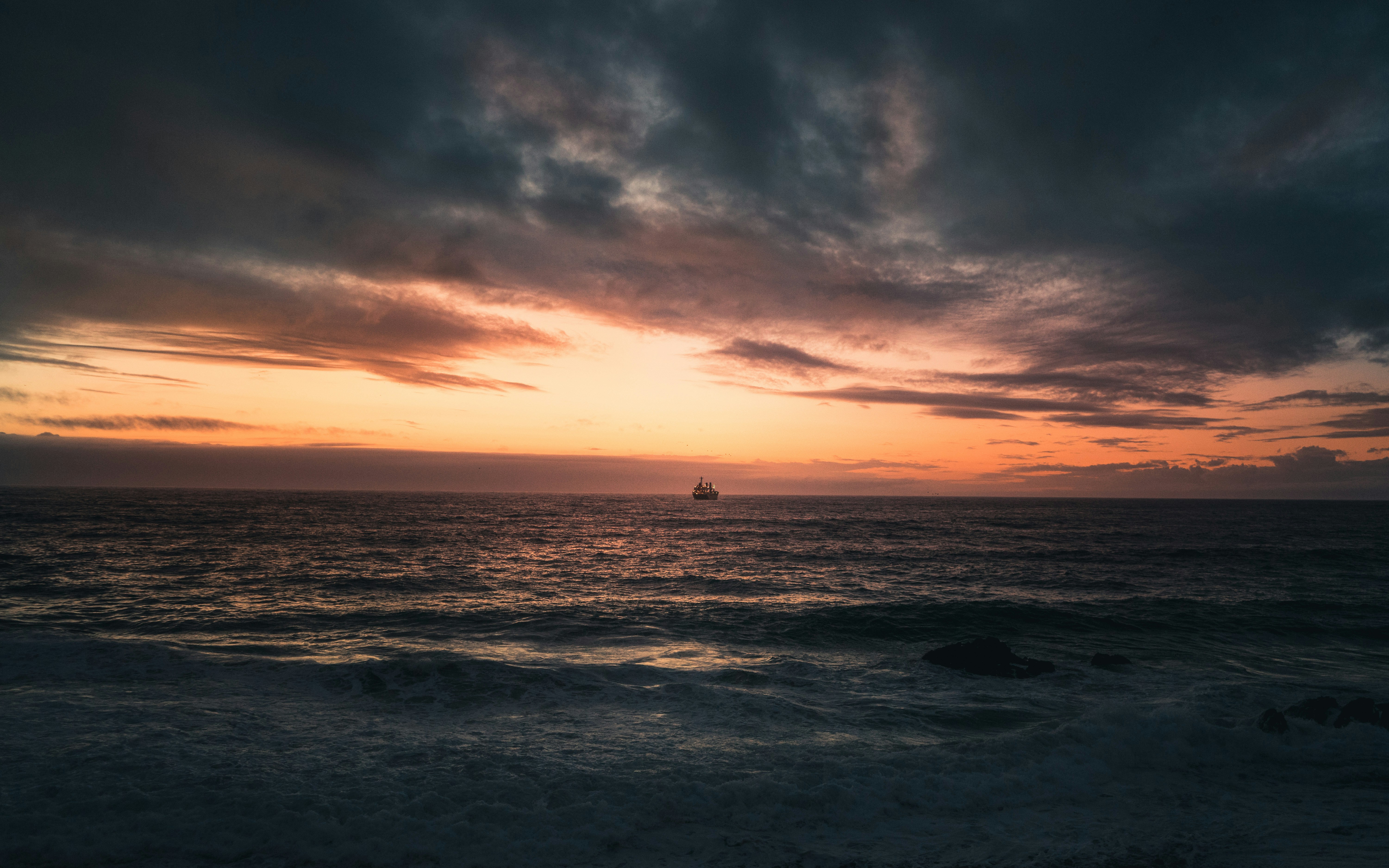 Vibrant sunset over a calm ocean with a distant silhouette of a boat under dramatic clouds.