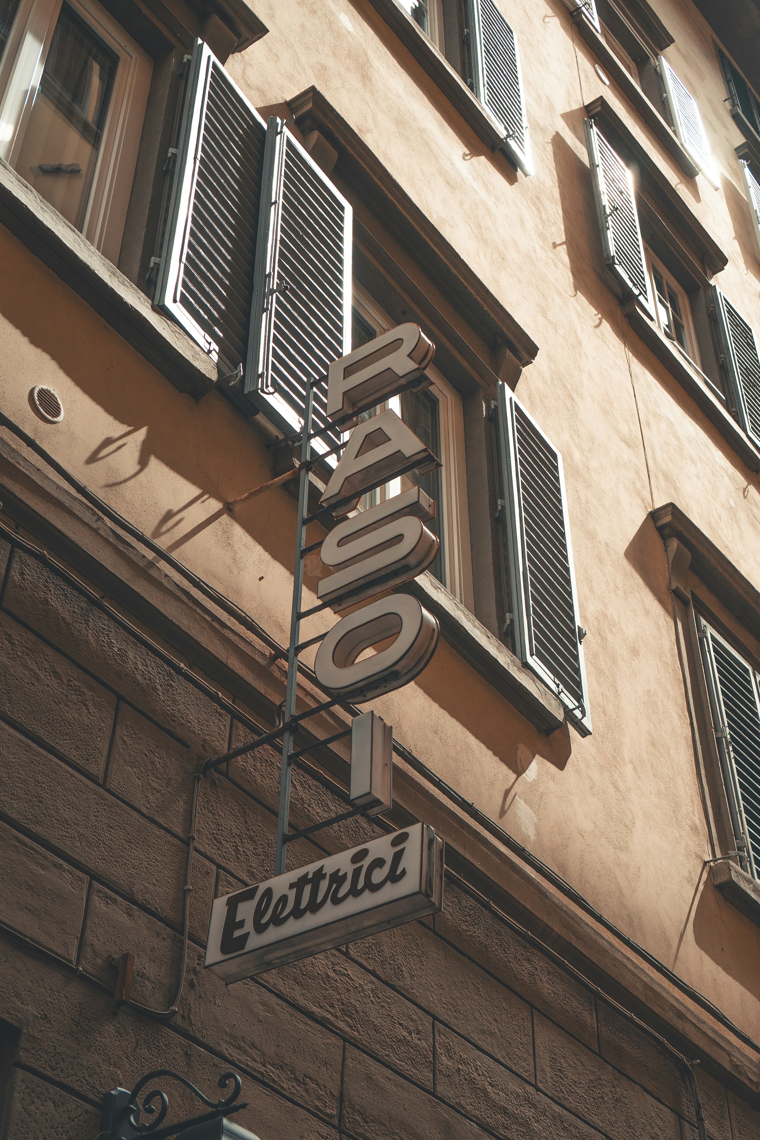 Vintage shop sign reading 'Raso Elettrici' against a textured wall, framed by classic window shutters.