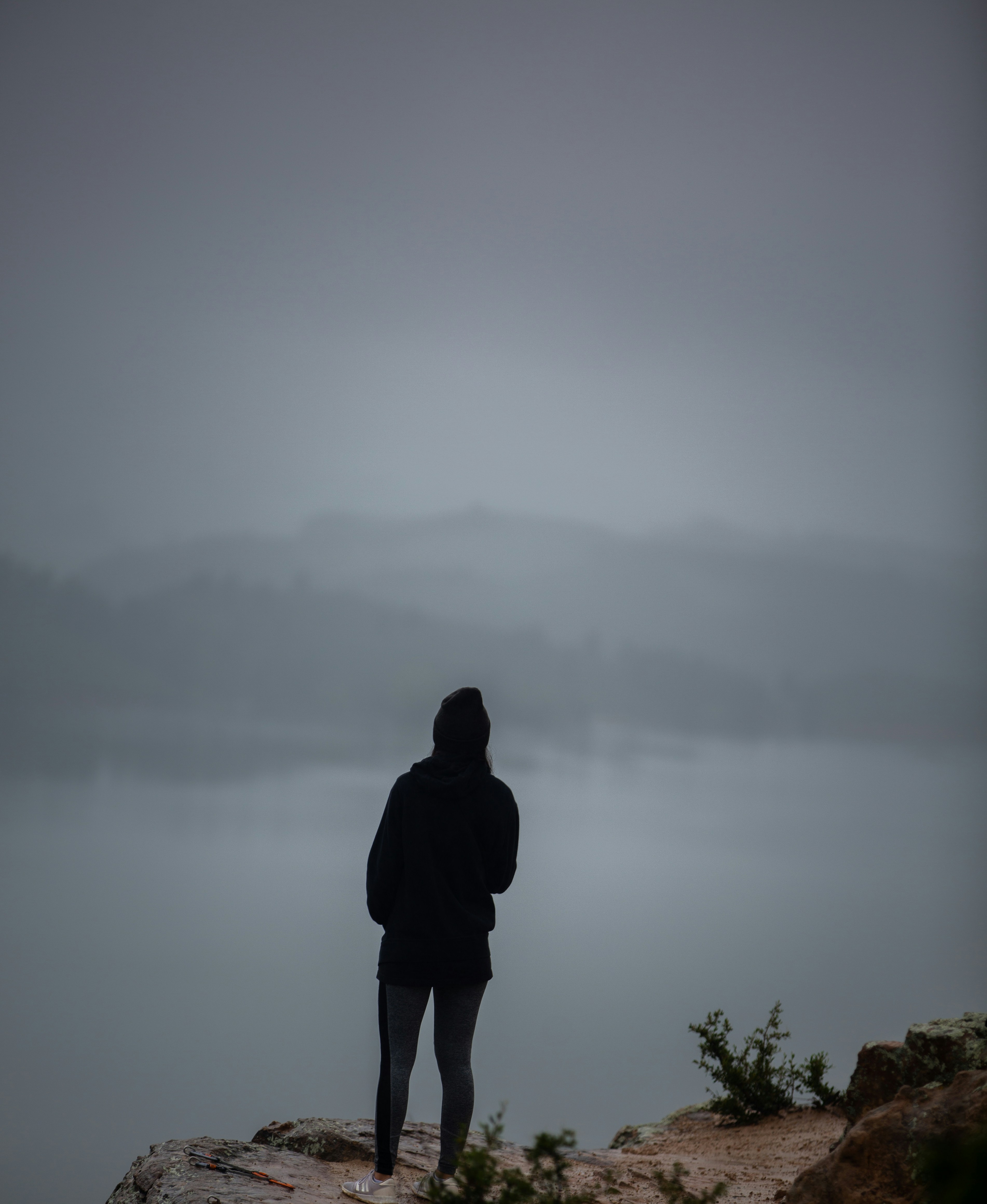 Person in a black hoodie stands on a rocky shore, gazing at the misty lake and distant hills. The atmosphere evokes a sense of solitude and contemplation.