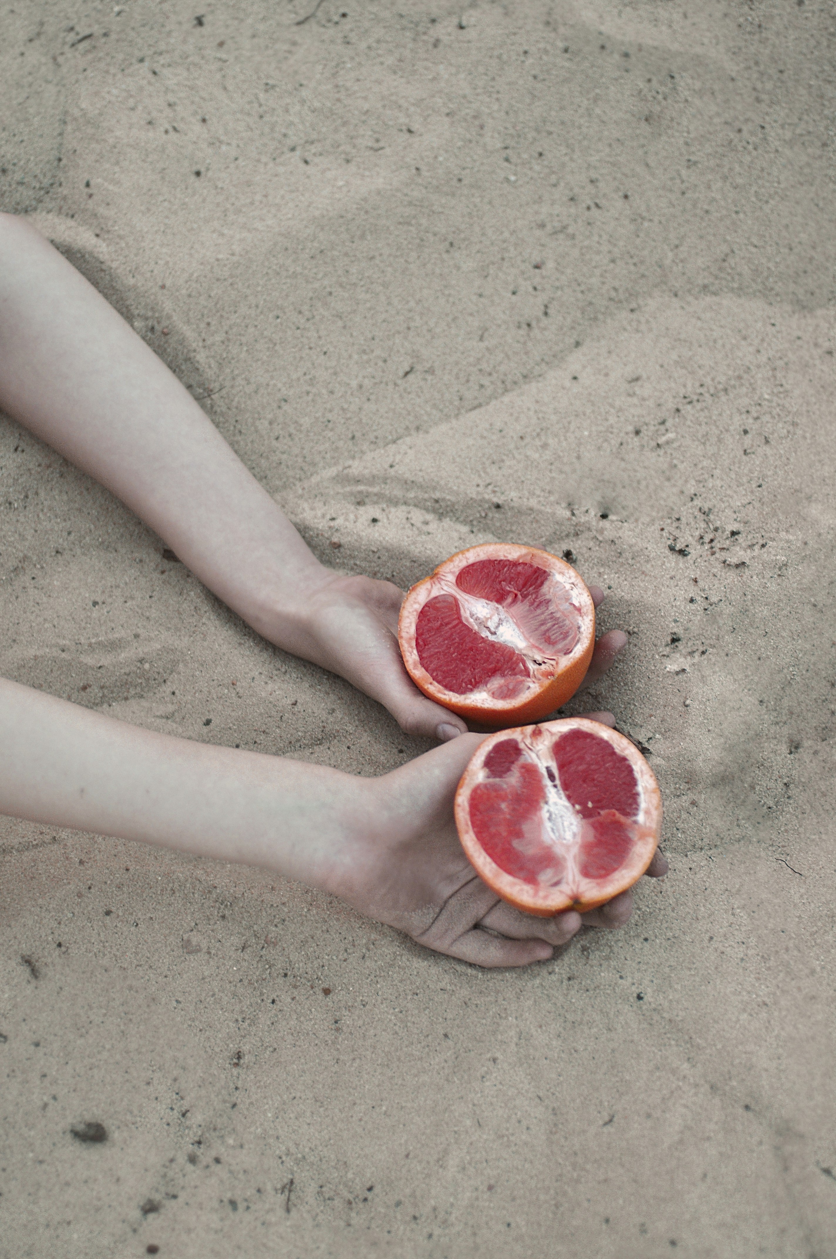 Two halves of a grapefruit held in hands, resting on a sandy surface. The earthy tones create a contrast with the vibrant fruit.