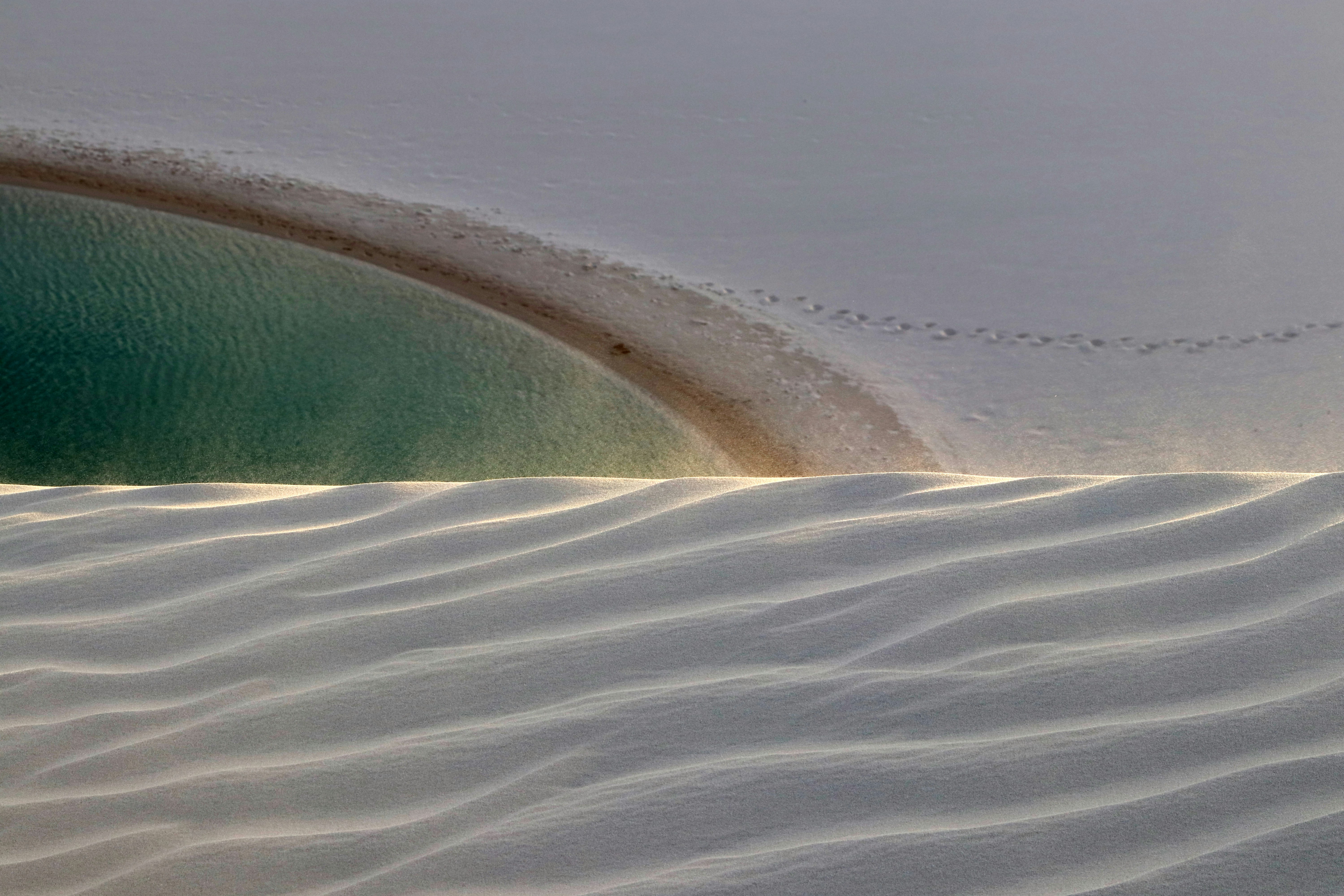 Gentle curves of sand meet a tranquil body of water, with subtle footprints tracing the shoreline. The interplay of textures creates a harmonious landscape.
