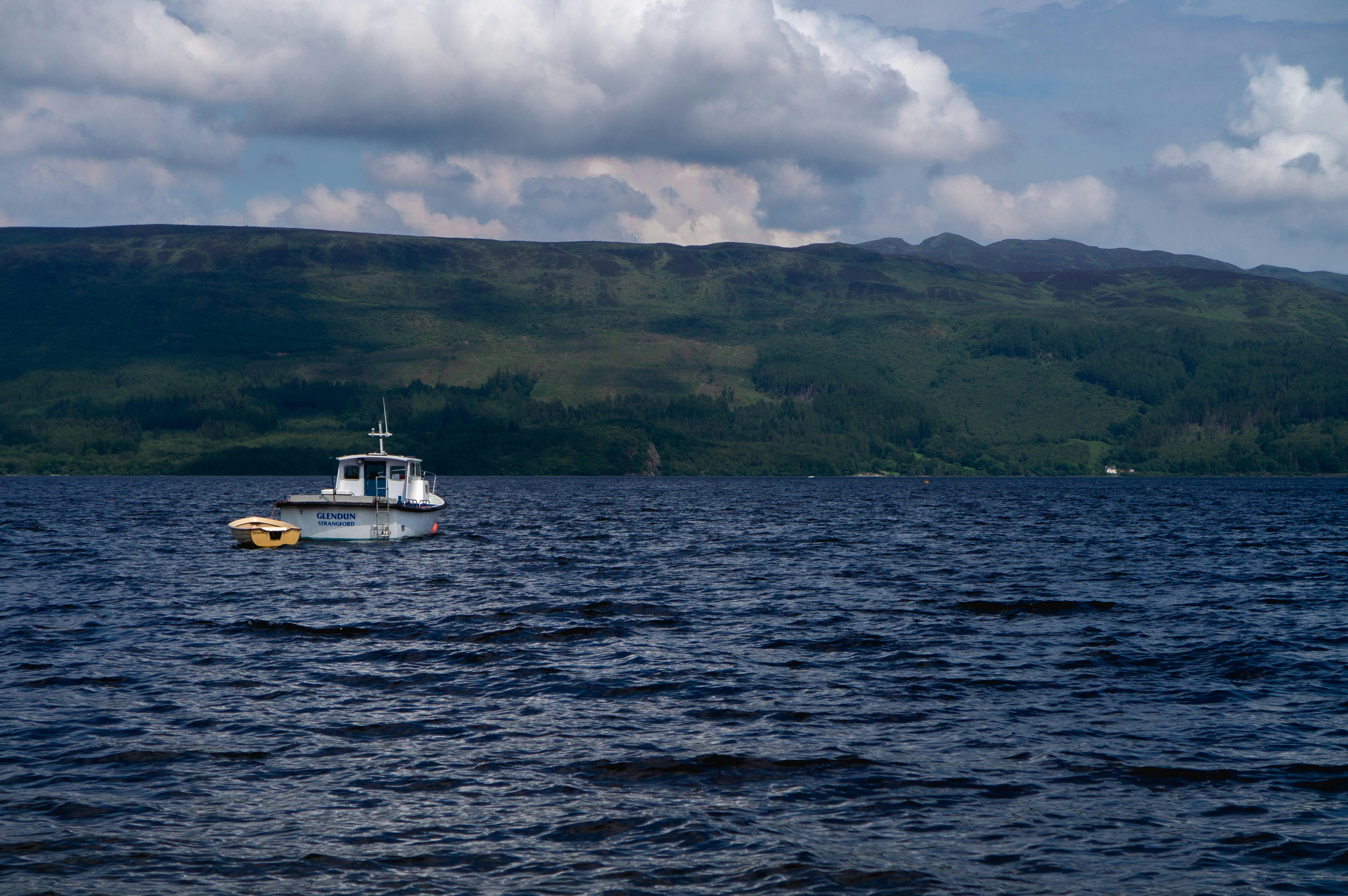 A solitary boat floats on the rippling waters of Loch Ness, framed by lush, green hills under a partly cloudy sky.