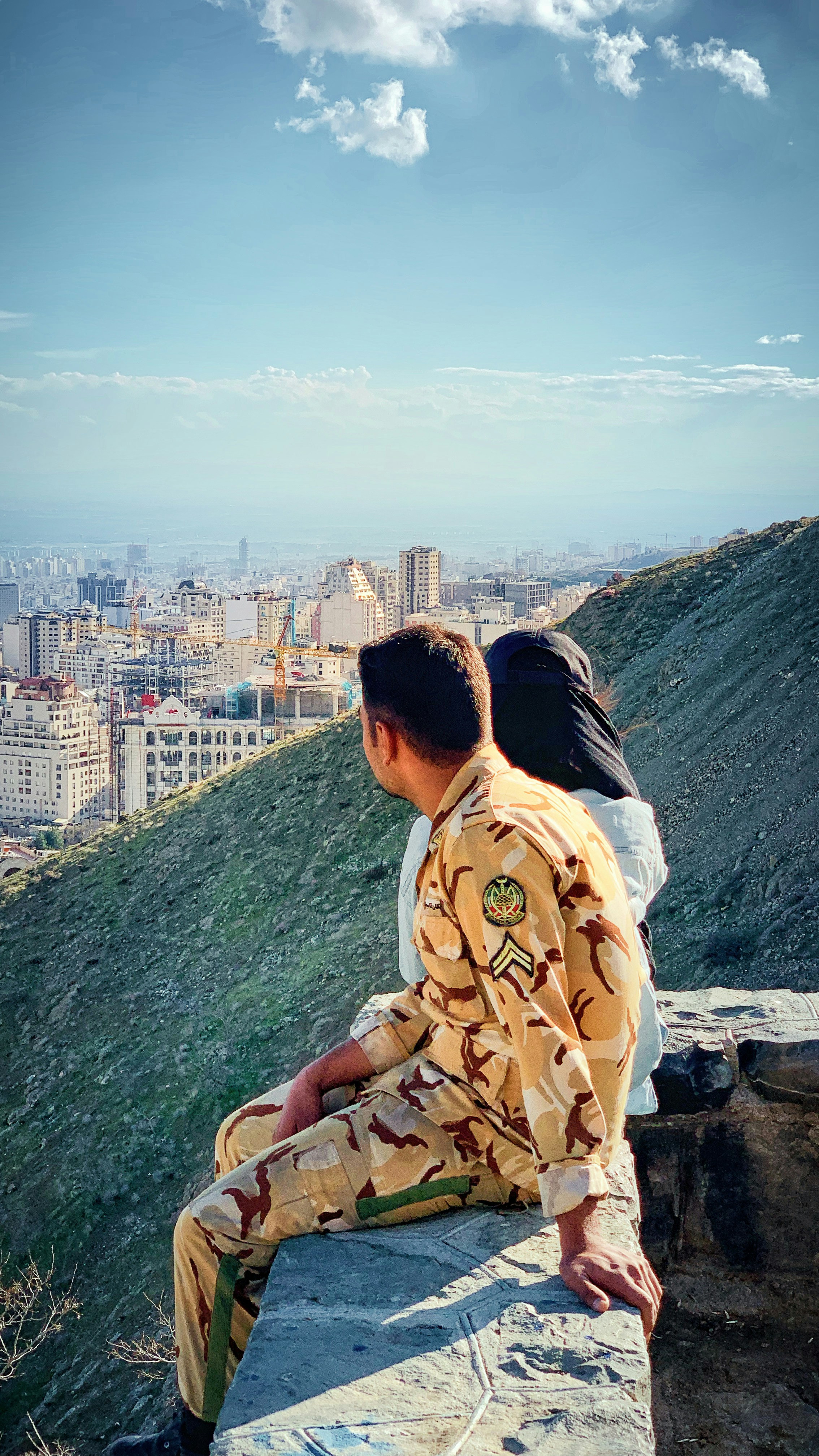 Two individuals seated on a rocky outcrop, overlooking a sprawling cityscape with a backdrop of distant mountains. The scene captures a moment of reflection amidst urban life.