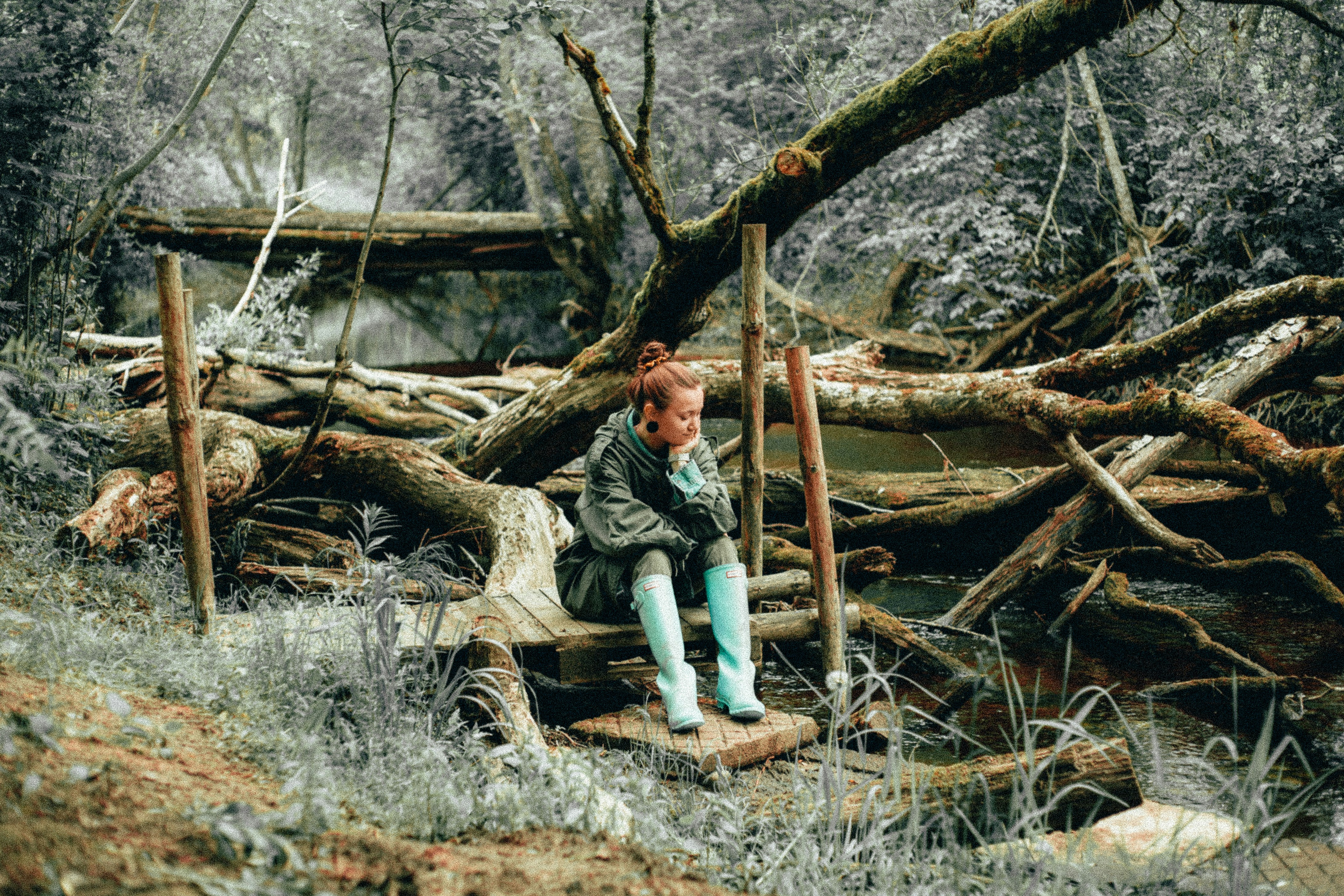 A person in green attire sits pensively on a wooden dock surrounded by fallen trees and lush greenery, evoking a sense of tranquility. The scene captures the harmony between humanity and nature.