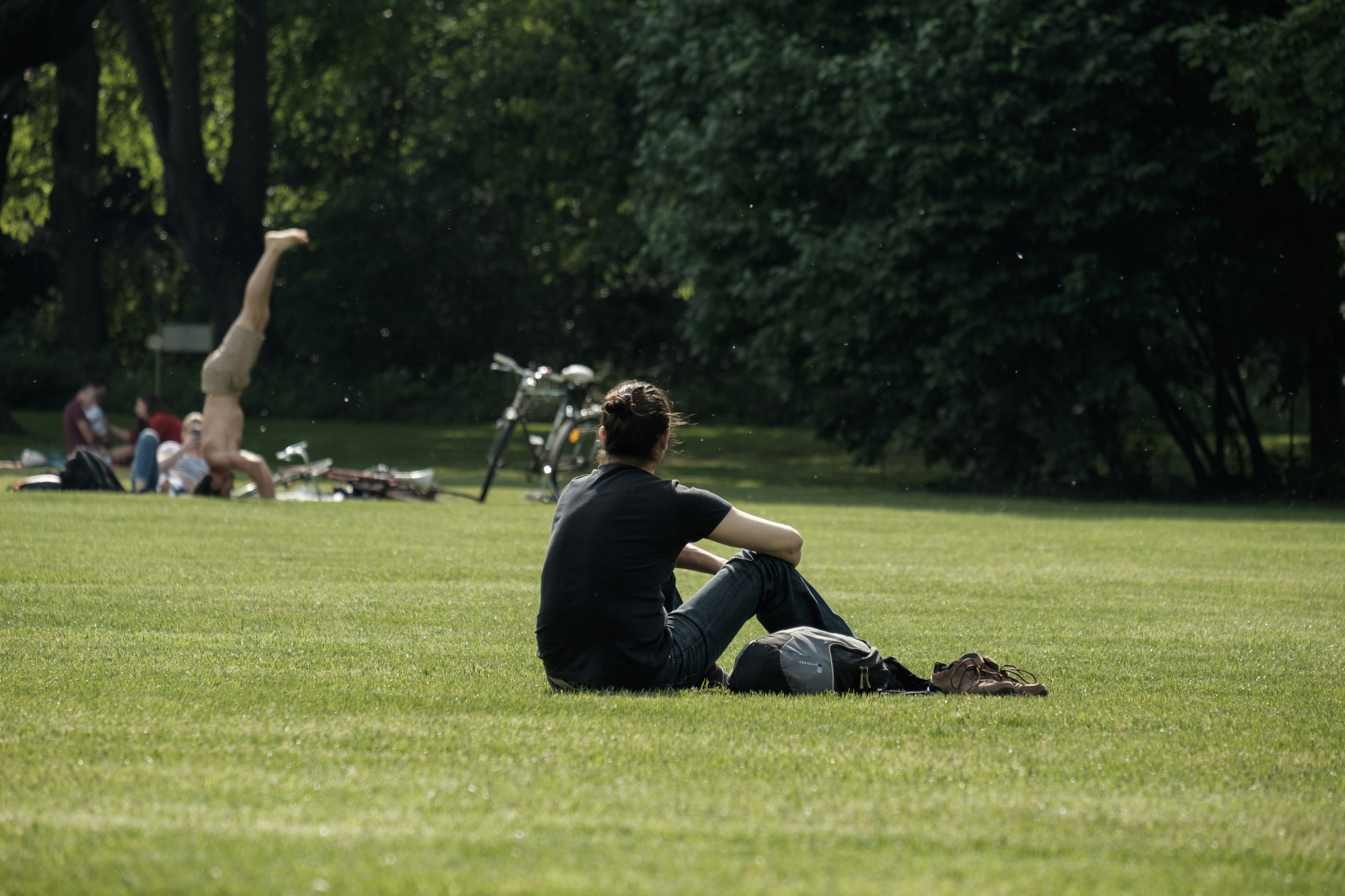 Photograph of a man sitting on a sunlit park lawn with a backpack beside him, while a bicycle and a few people relax in the background.