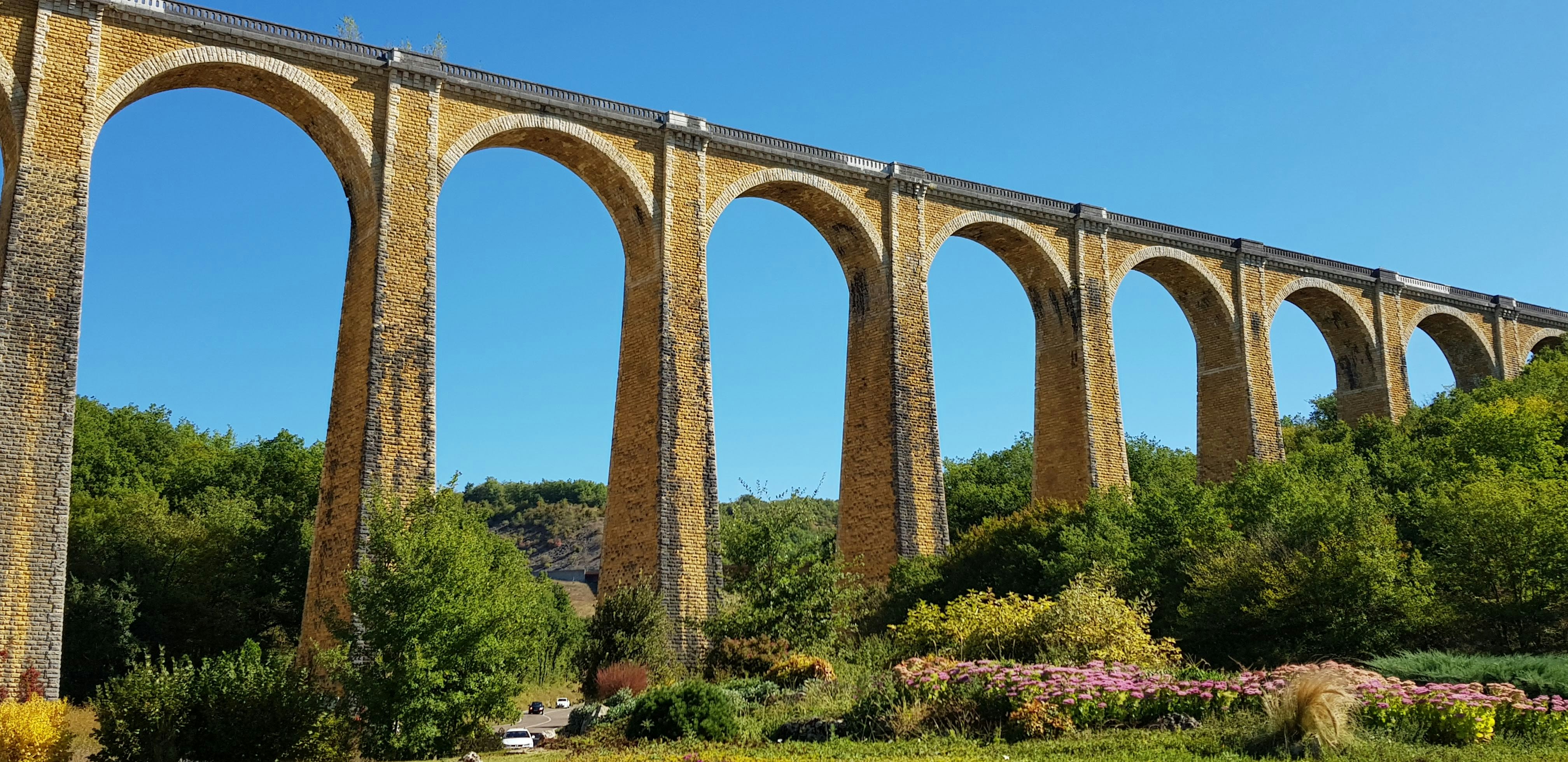 Historic viaduct rises majestically above lush greenery, framed by a clear blue sky. The vibrant flora below adds a splash of color to the scene.
