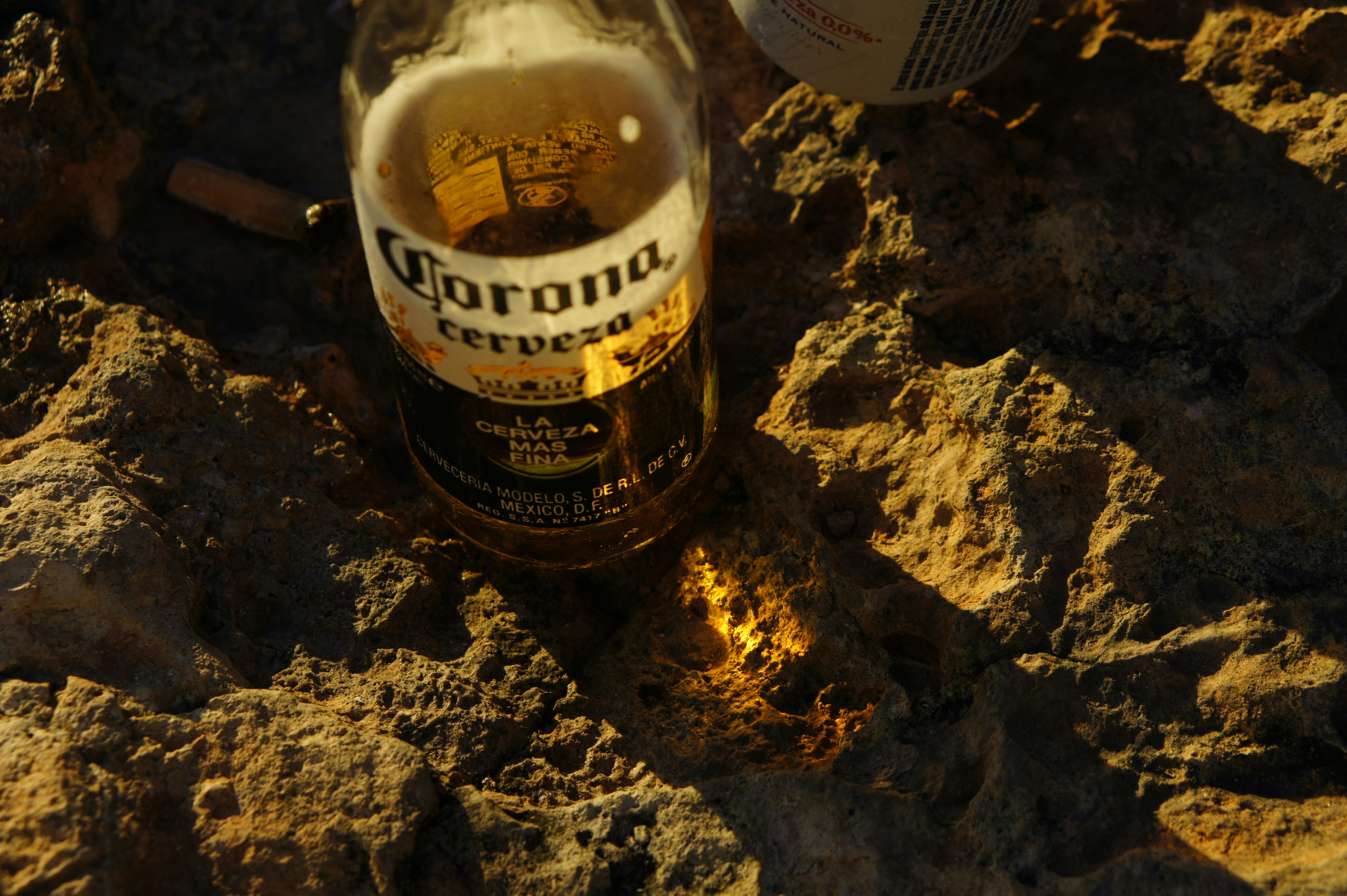 A Corona beer bottle resting on rugged terrain, casting reflections on the textured rocks beneath. The warm light enhances the scene's natural charm.