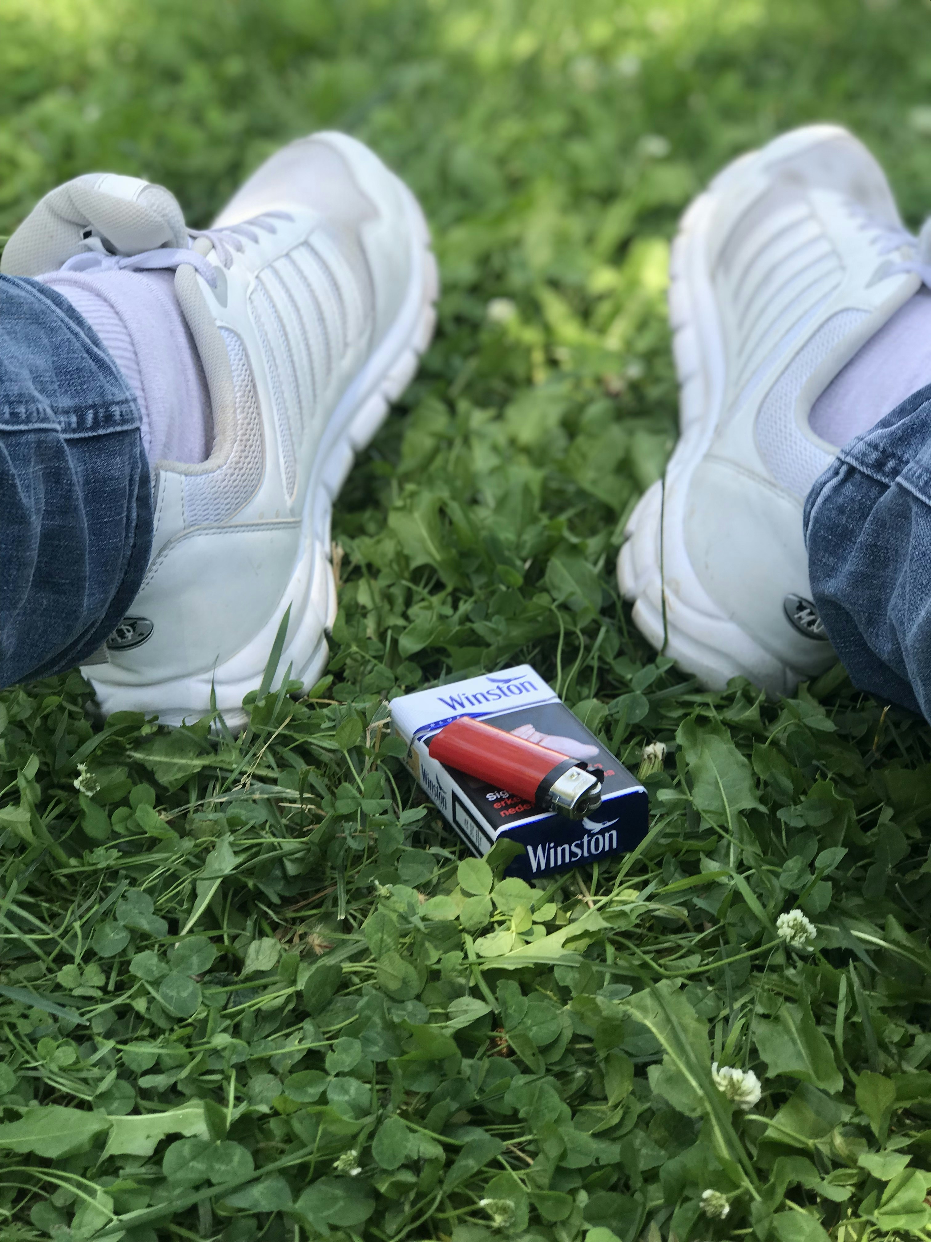 person in blue denim jeans and pair of white Nike sneakers sitting on grasses with black and white Winston cigarettes pack