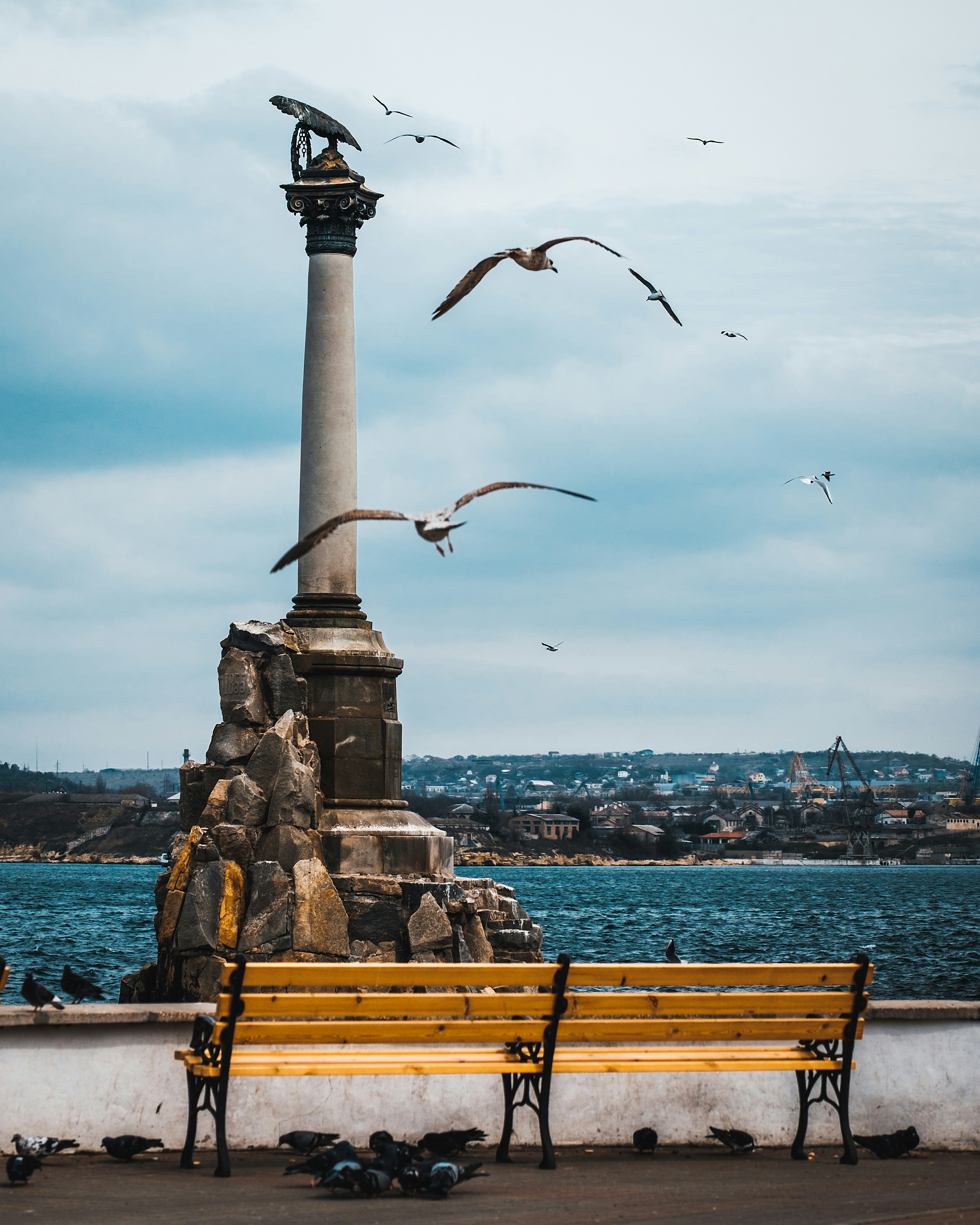 A tall column with a statue stands prominently on the rocky shore, surrounded by flying birds and a serene coastal backdrop. A yellow bench invites contemplation nearby.