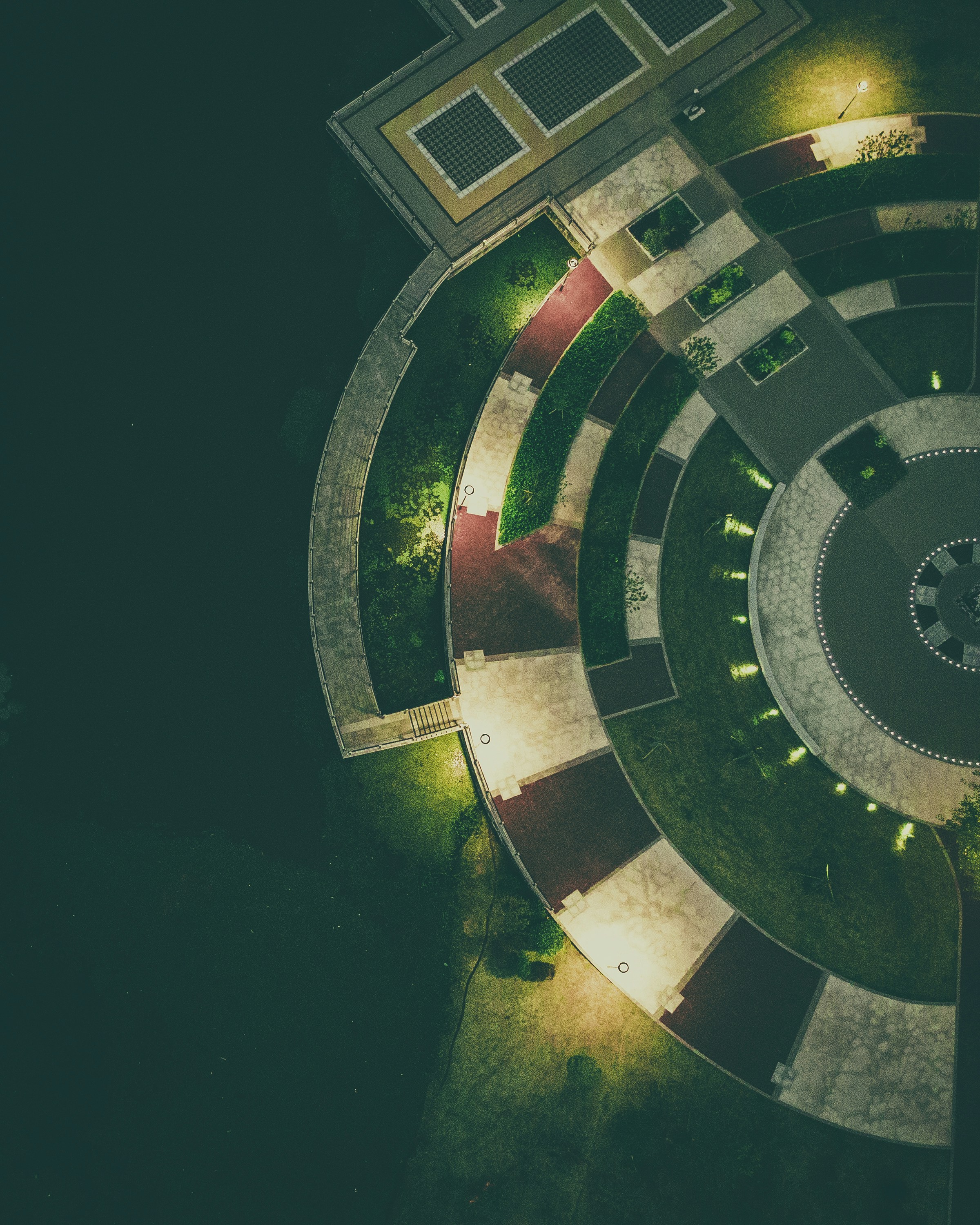 Aerial view of a beautifully designed circular park illuminated at night, showcasing pathways and greenery. The layout emphasizes geometric patterns and light contrasts.