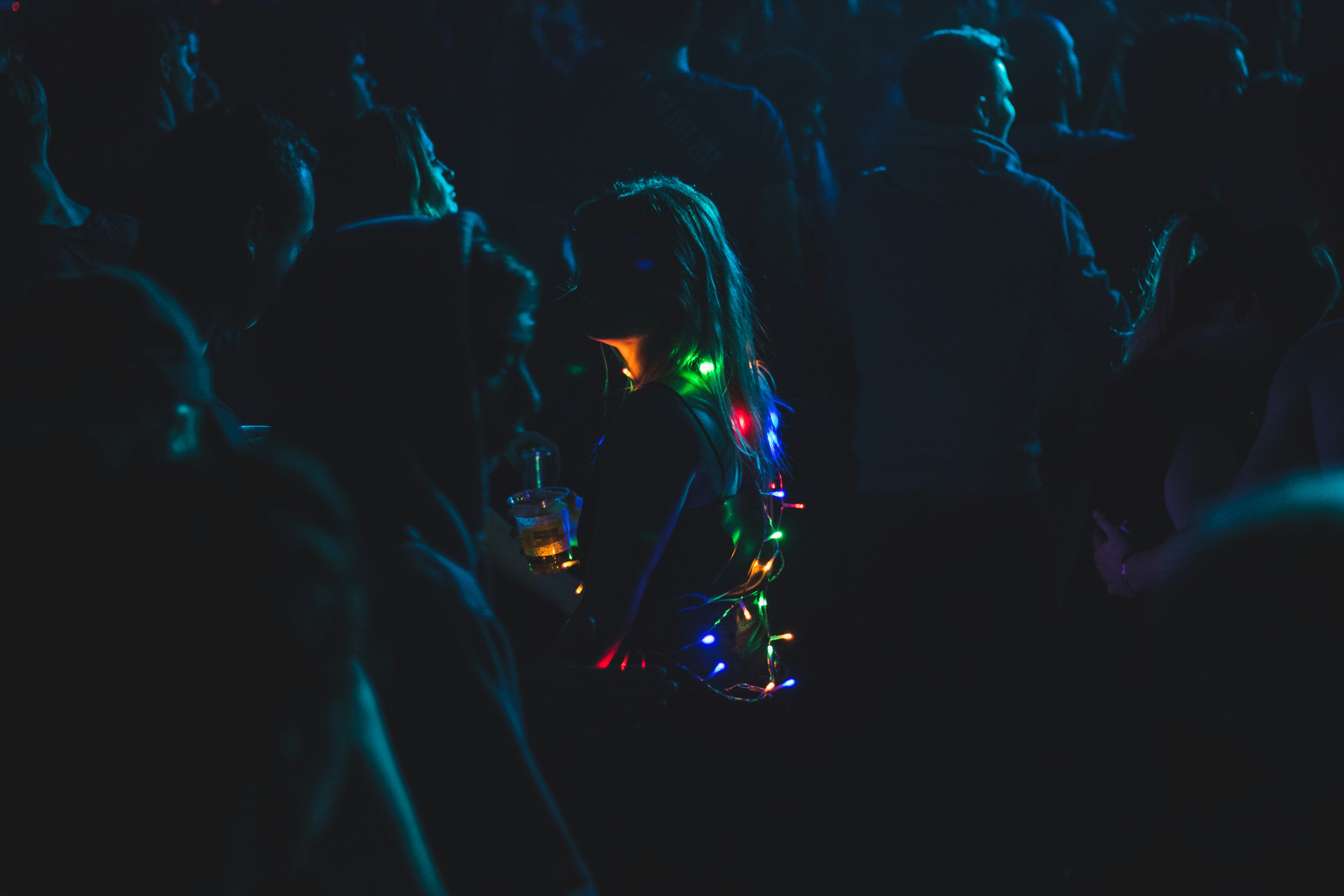 A partygoer adorned with colorful lights stands amidst a crowd, creating a striking contrast against the dark surroundings.