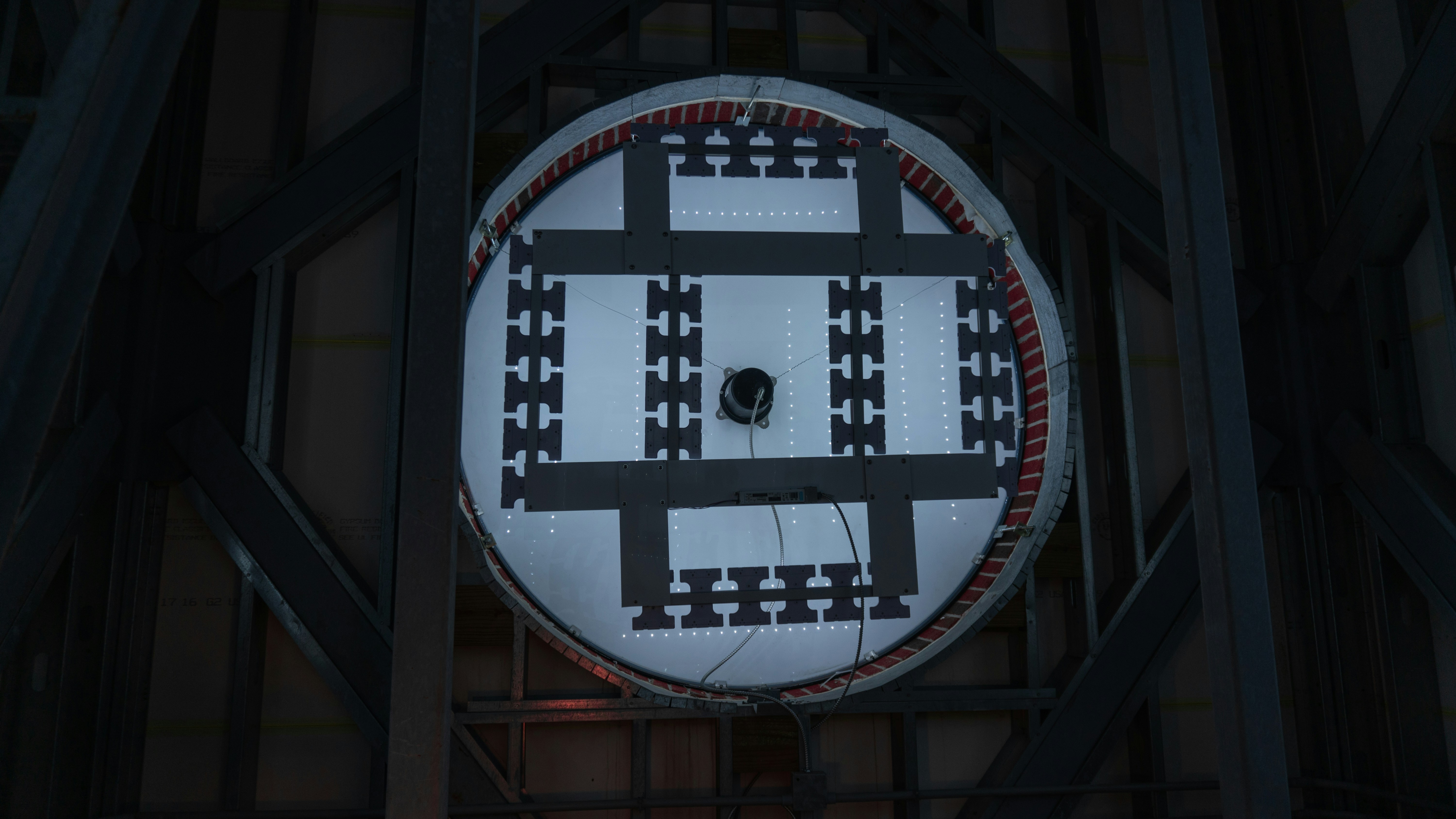 Circular engineering component suspended in a structure, showcasing intricate design and technology. Illuminated from below, emphasizing its geometric features.