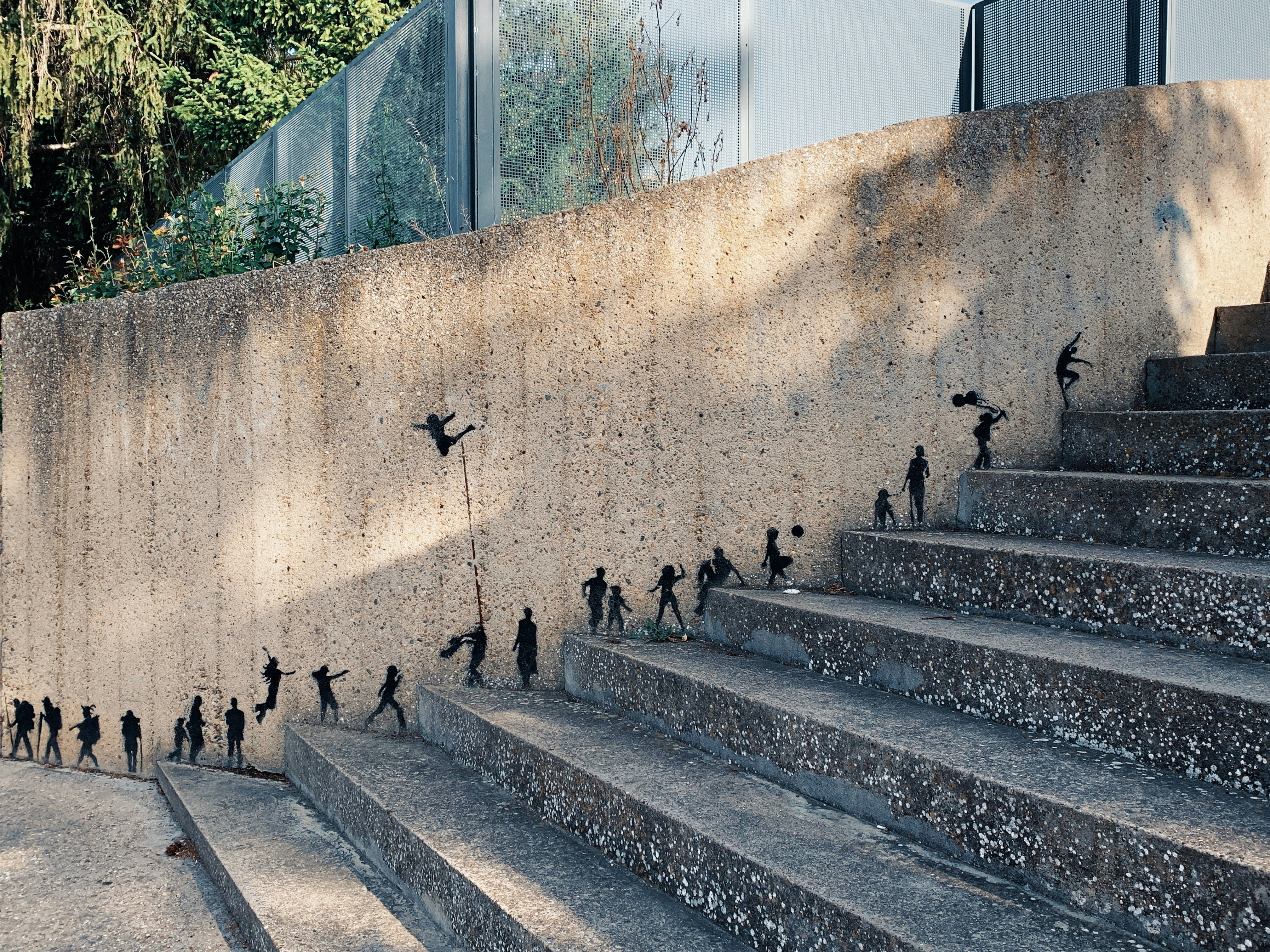 Line of black silhouette figures climbs a stepped, textured concrete wall beside a glass barrier.