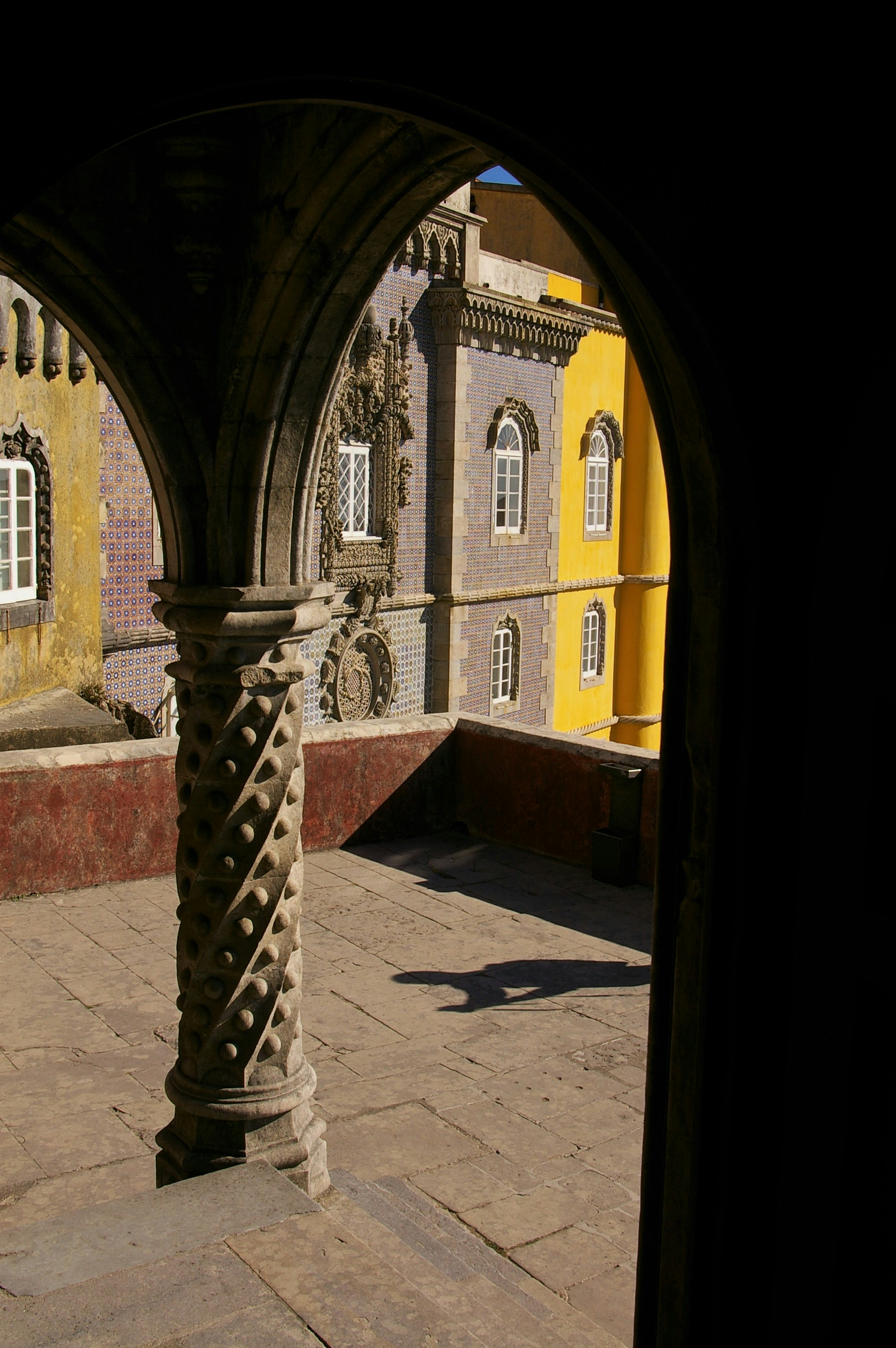 Intricate stone column framed by an arch, revealing vibrant buildings in the background. The play of light and shadow adds depth to the scene.