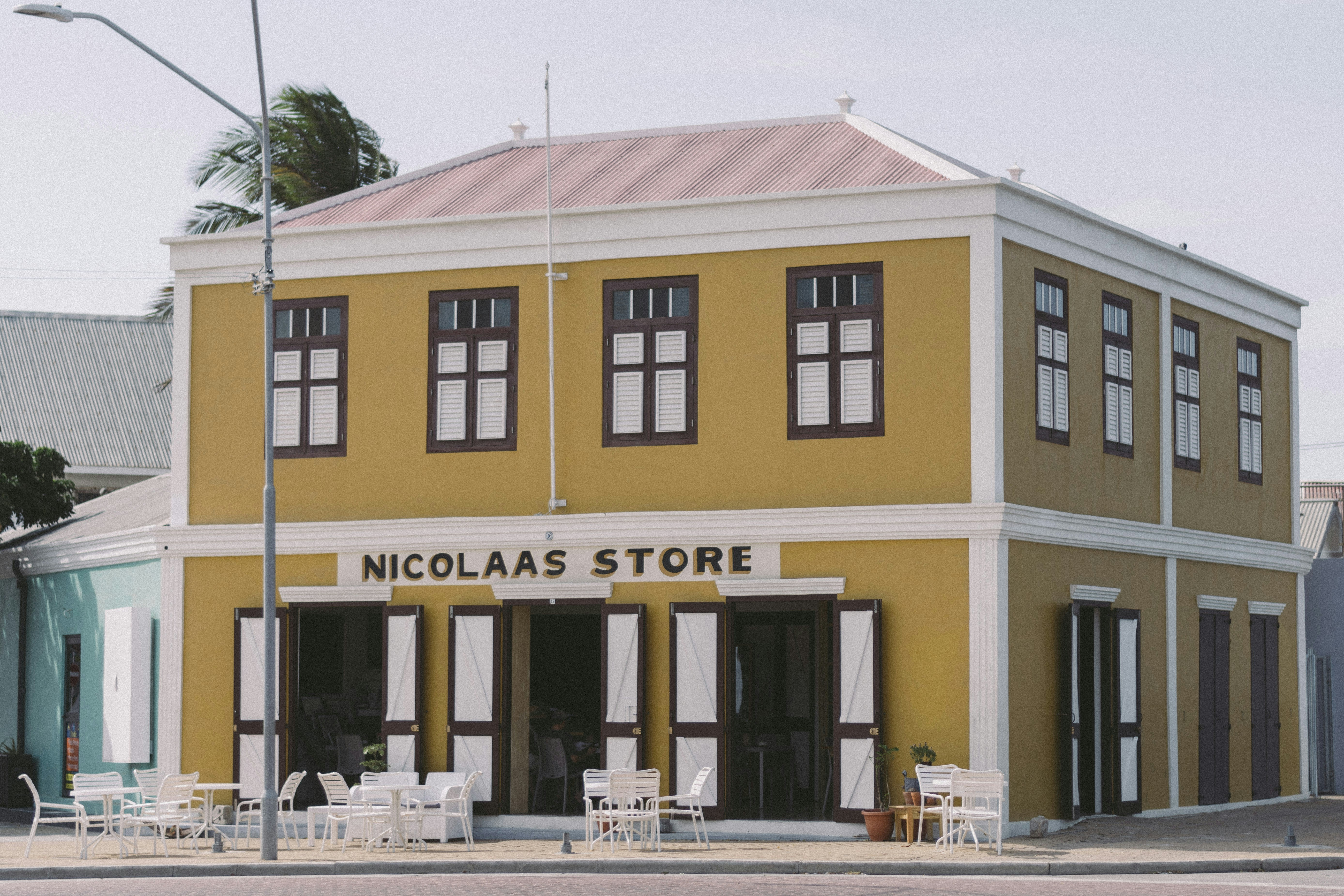 Two-story yellow building with white trim and a red roof, labeled 'Nicolaas Store,' featuring outdoor seating under clear skies.