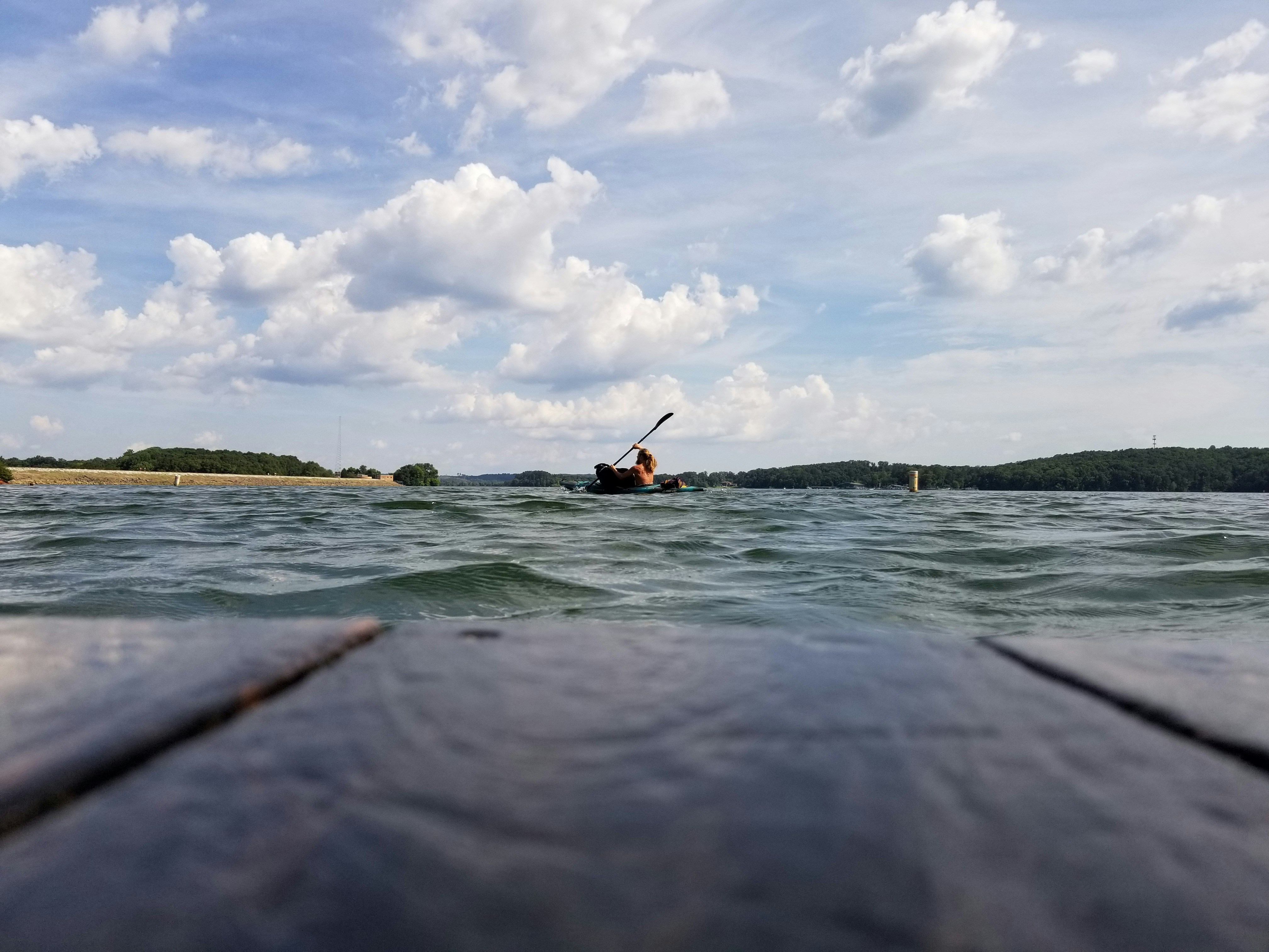 A kayaker glides across serene waters under a sky dotted with fluffy clouds, framed by distant hills. The perspective emphasizes the calmness of the scene.
