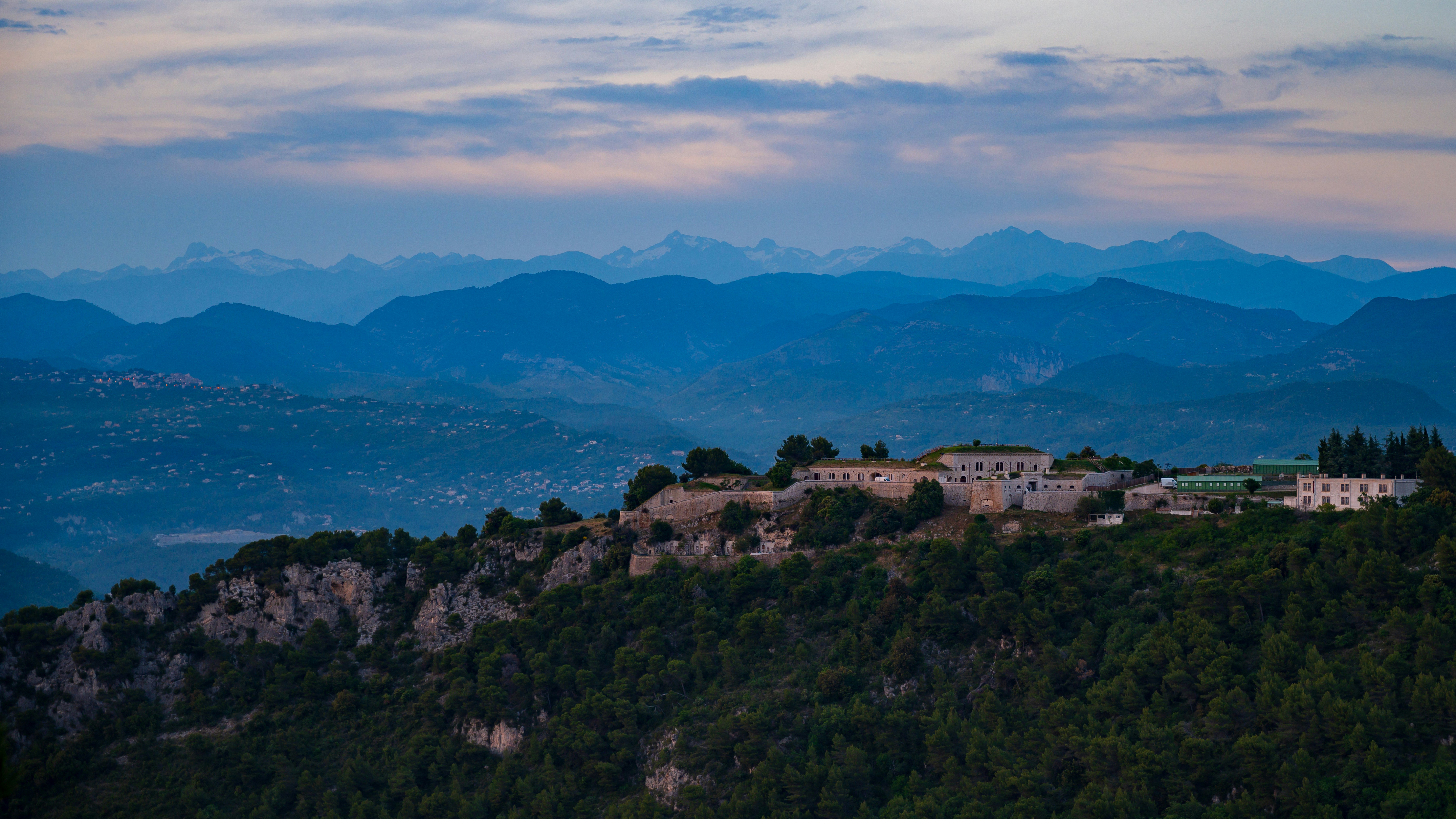 Historic fortress perched on a lush cliff with layers of blue mountains in the background at dusk.