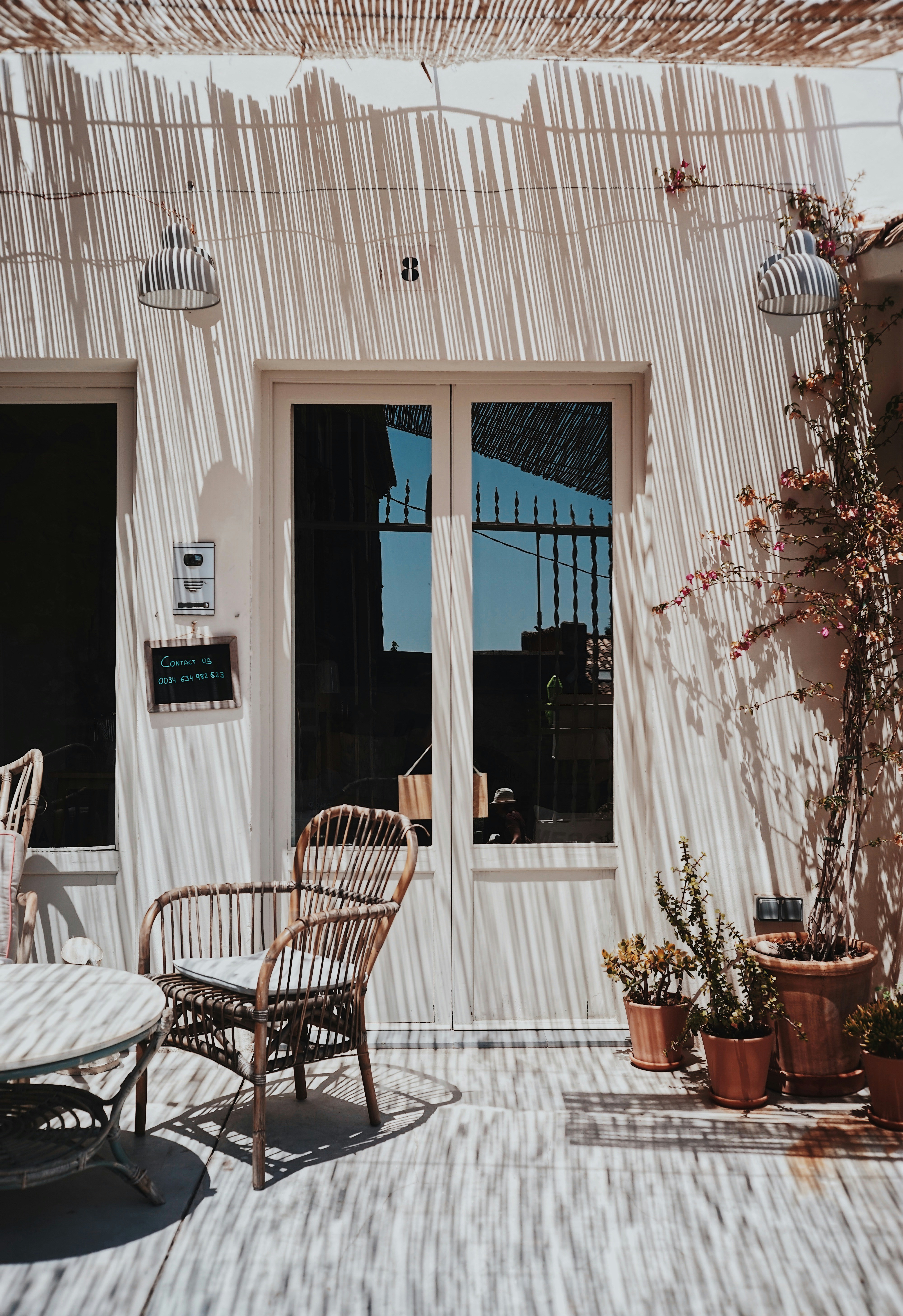 A sunlit patio featuring a wicker chair and table, adorned with plants, casting intricate shadows on the walls. The open doors reflect a tranquil outdoor scene.