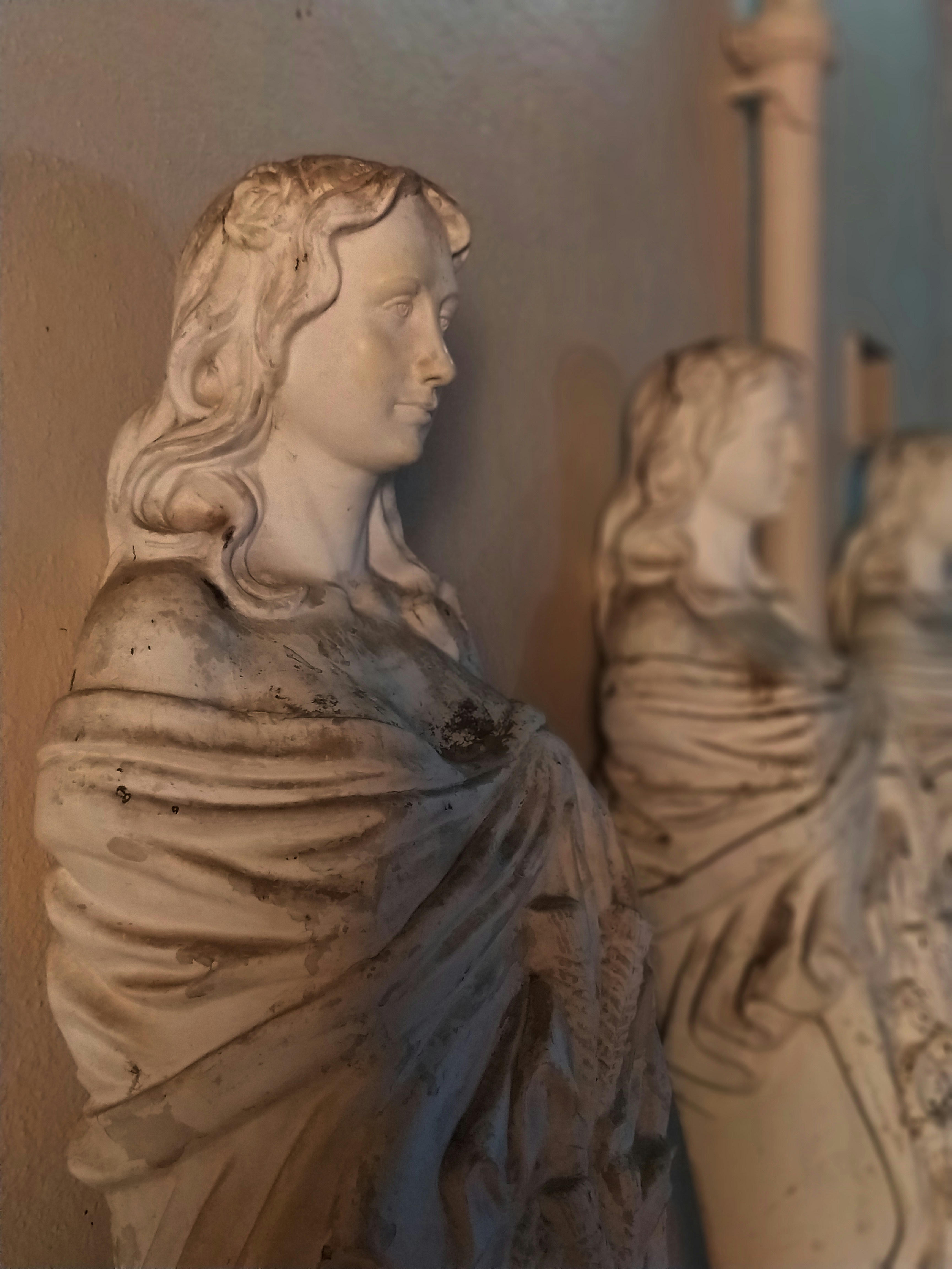 Three classical sculptures of women with flowing hair and draped garments, softly illuminated against a muted background.