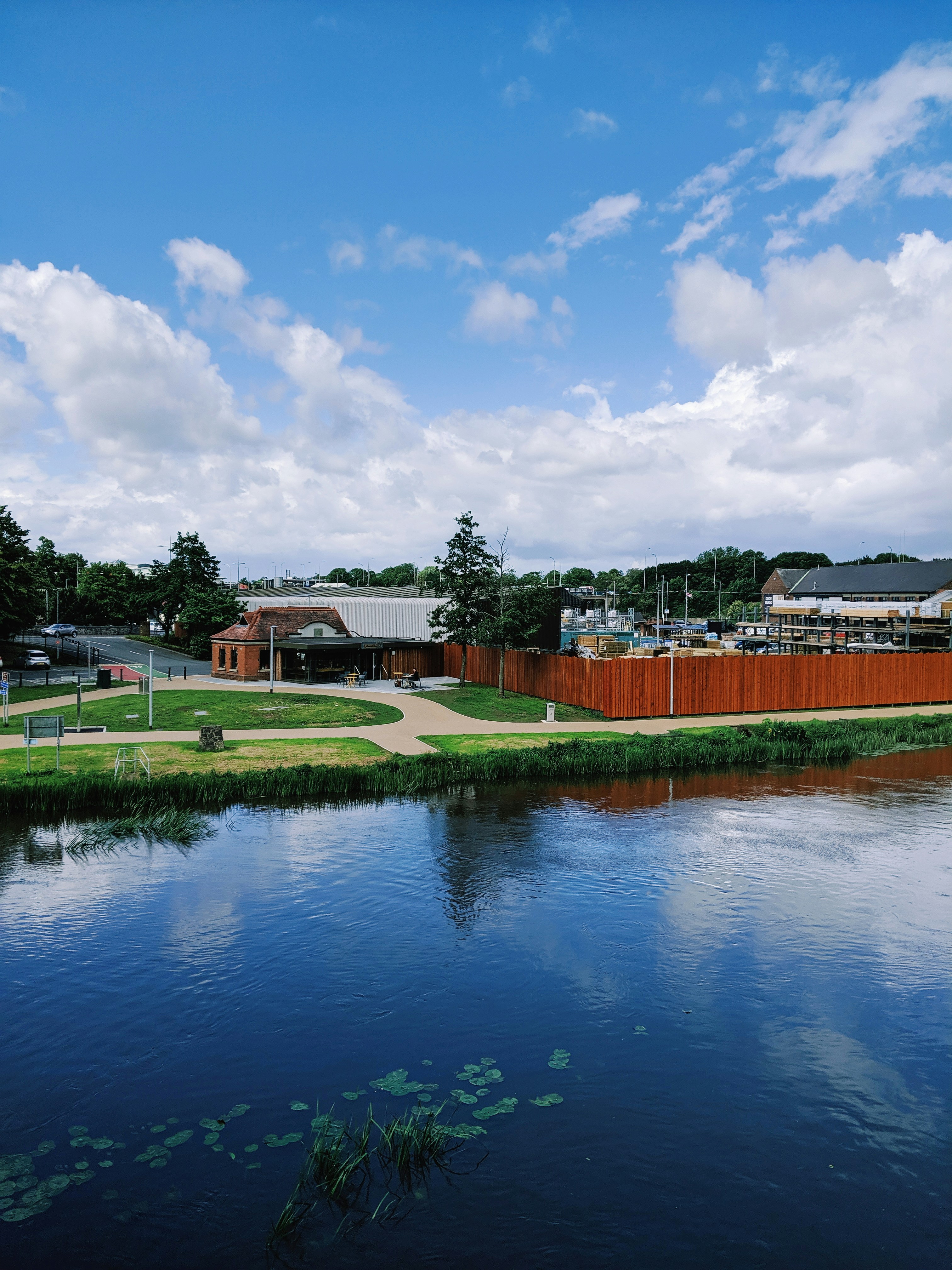 A serene riverside scene featuring a park, buildings, and vibrant greenery under a partly cloudy sky.